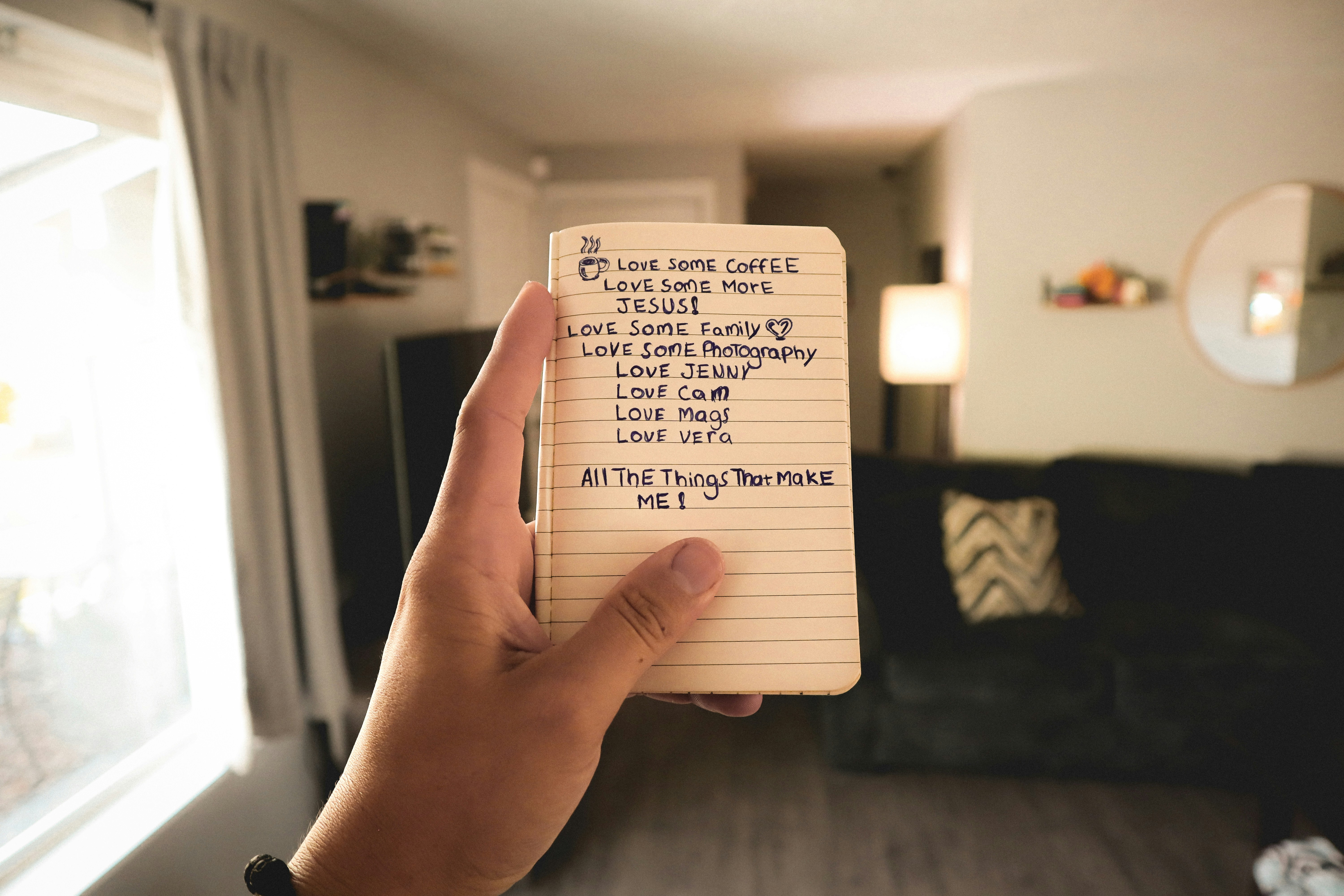 Close-up of a hand holding a small notebook with handwritten text, the cozy living room in the background. Note resembles a personal message board; the scene is warm and softly lit.