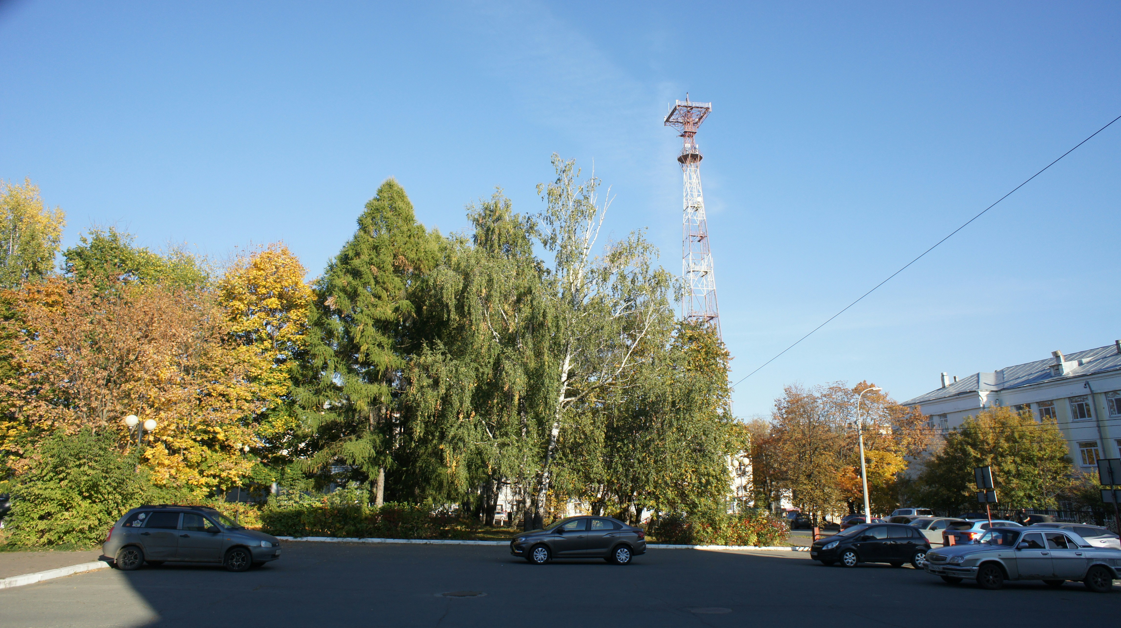 Wide street scene with parked cars and a distant tall radio mast among autumn trees, illustrating an urban edge landscape.