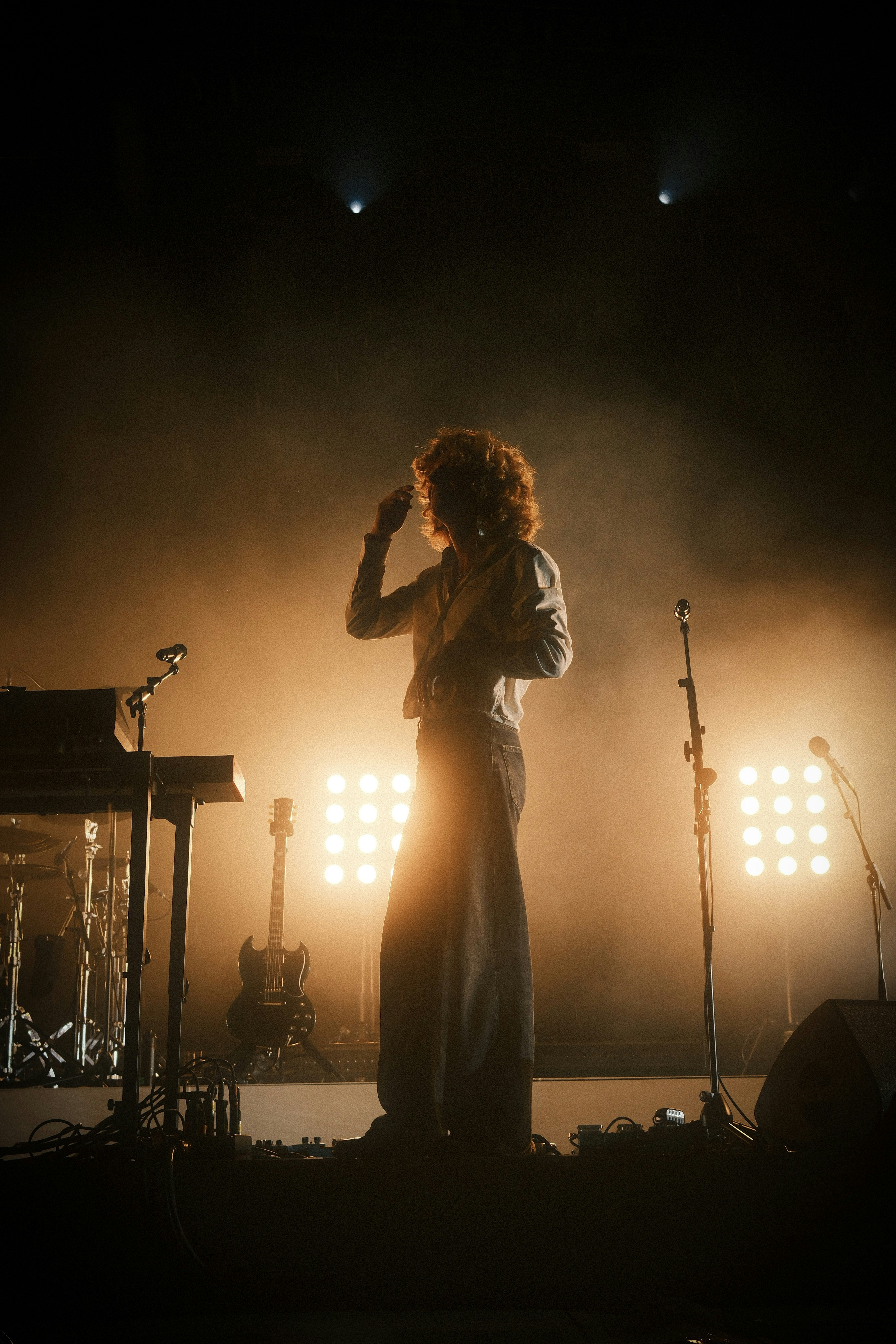 A lone performer stands center stage, backlit by warm amber lights and fog, with instruments and microphones flanking the scene.