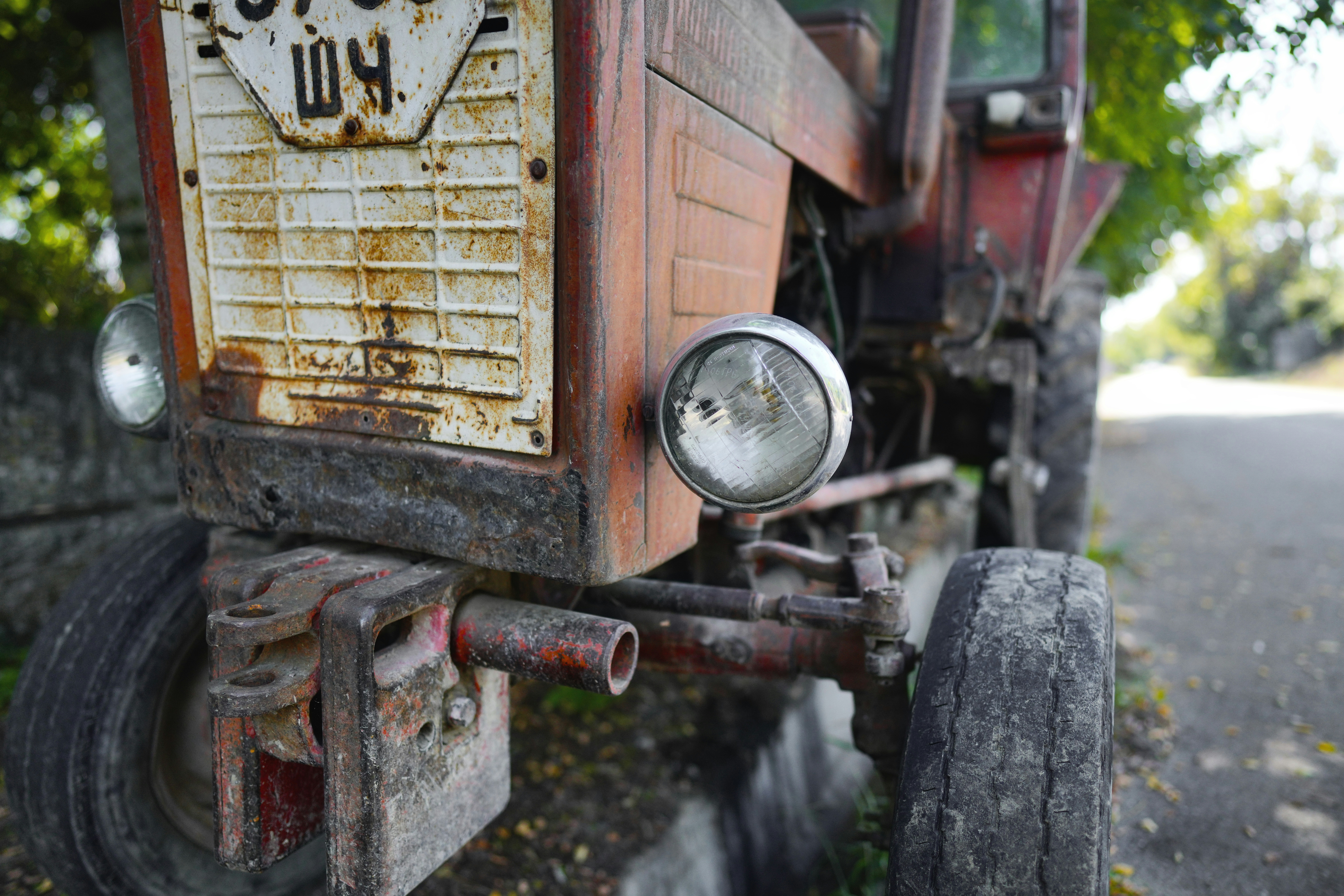 Close-up of a weathered orange tractor front, showing rust, a missing emblem, and a single round headlamp, resting on a dirt road with greenery in the background.
