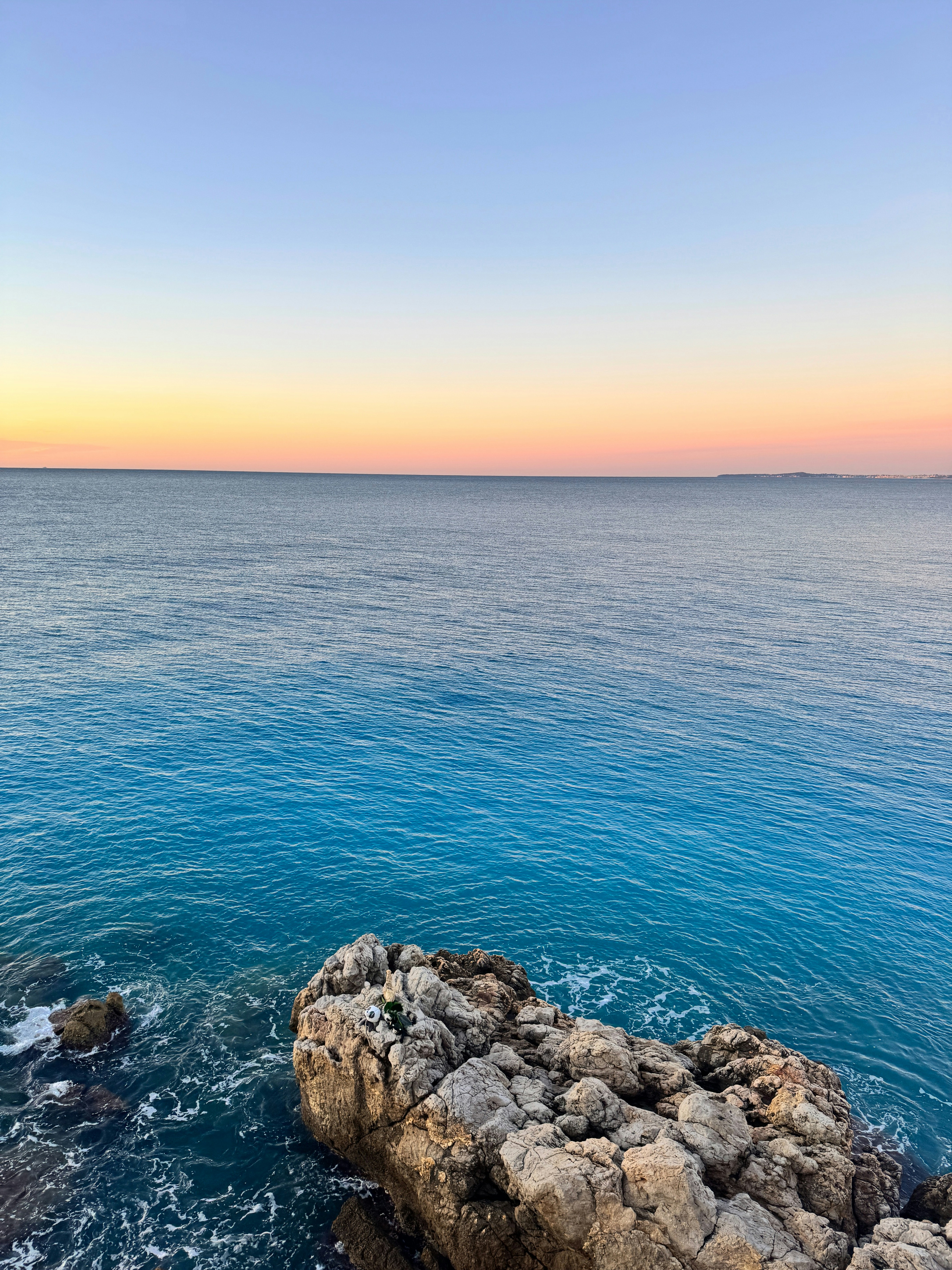 Coastline at sunset with calm blue waters and a rocky foreground, emphasizing vast horizon and tranquil mood. Photograph of a rocky outcrop meeting the sea at dusk.