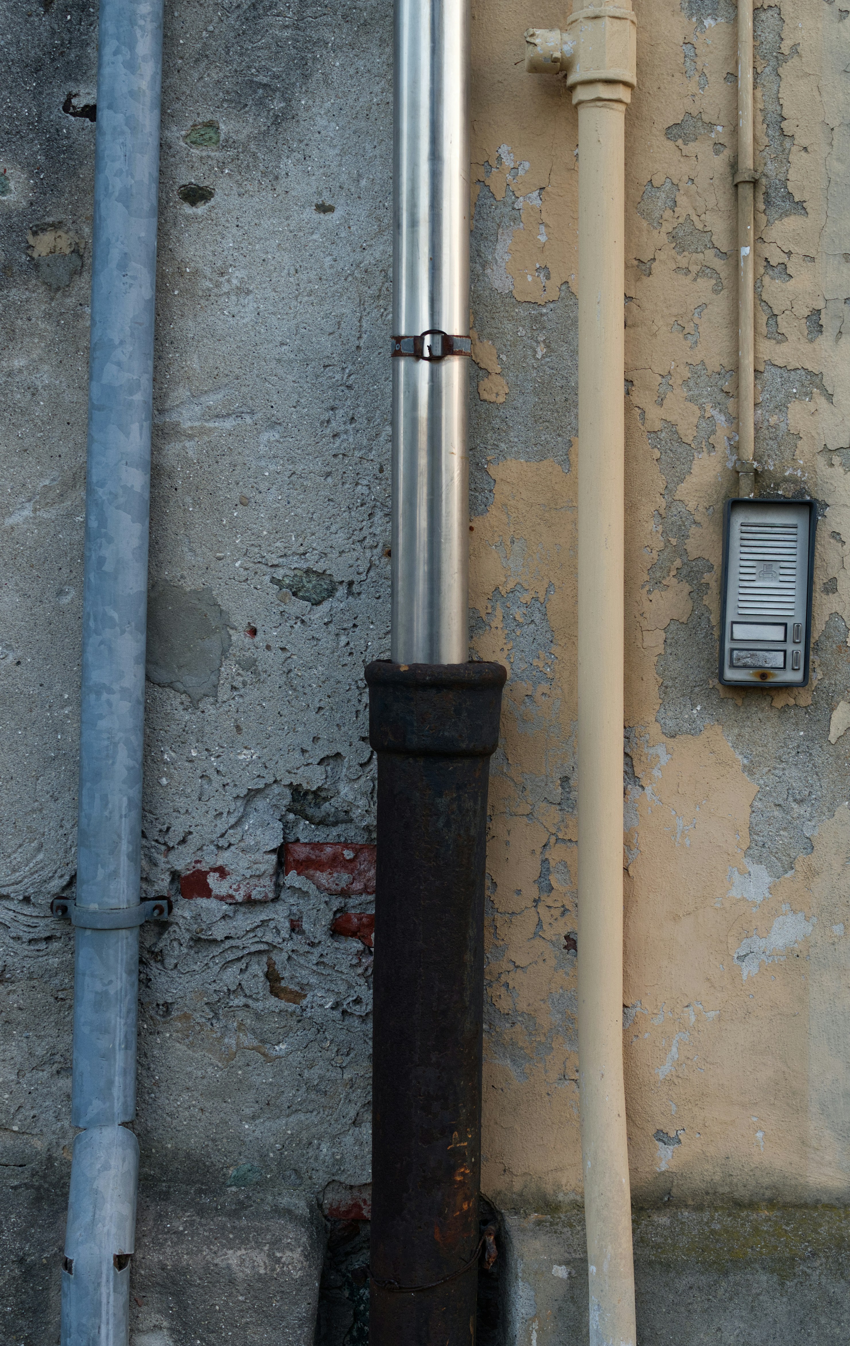 Three vertical pipes and an intercom on a weathered concrete and brick wall