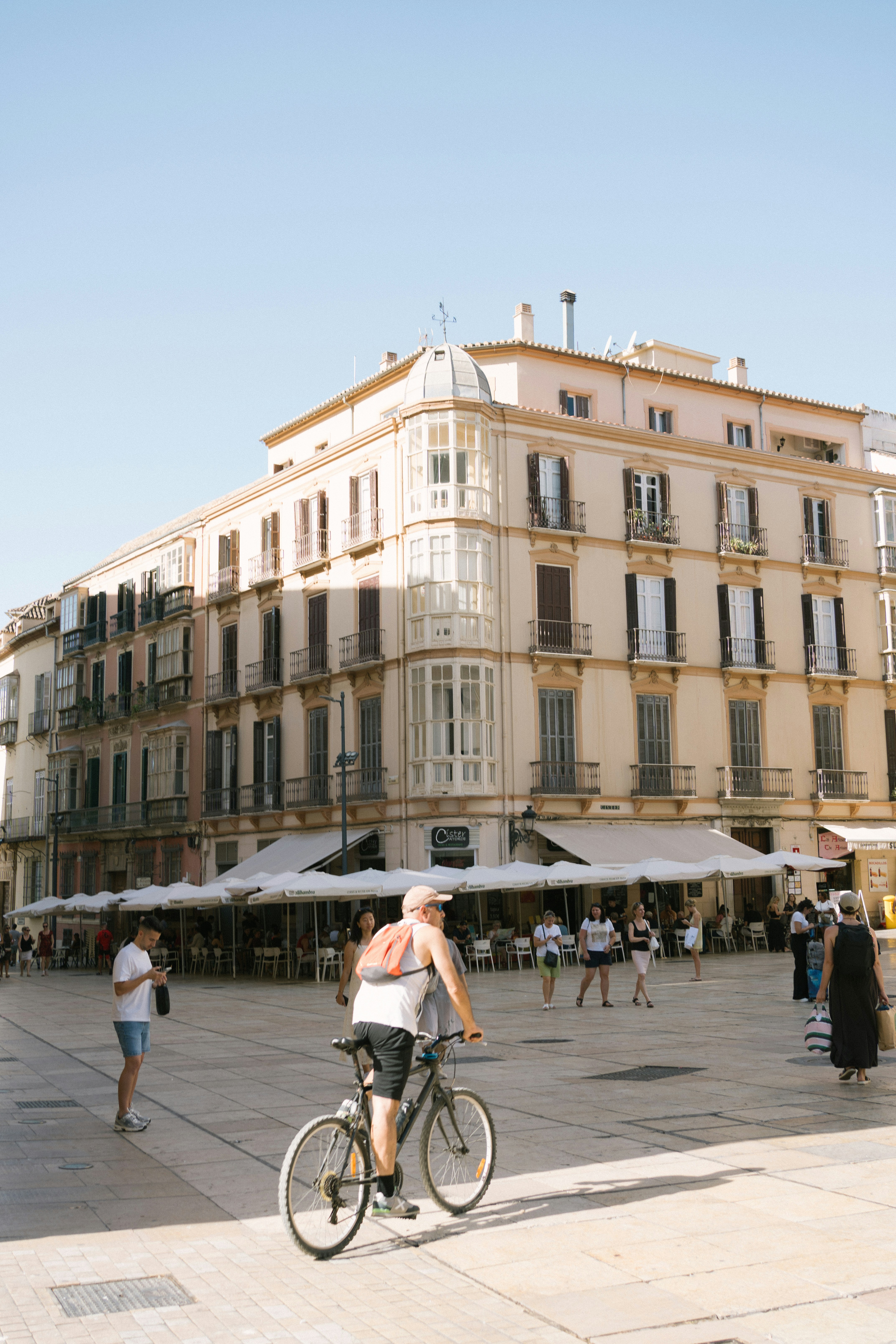 City square with a pale historic building under a clear blue sky; a cyclist rides by while pedestrians gather near outdoor cafes.