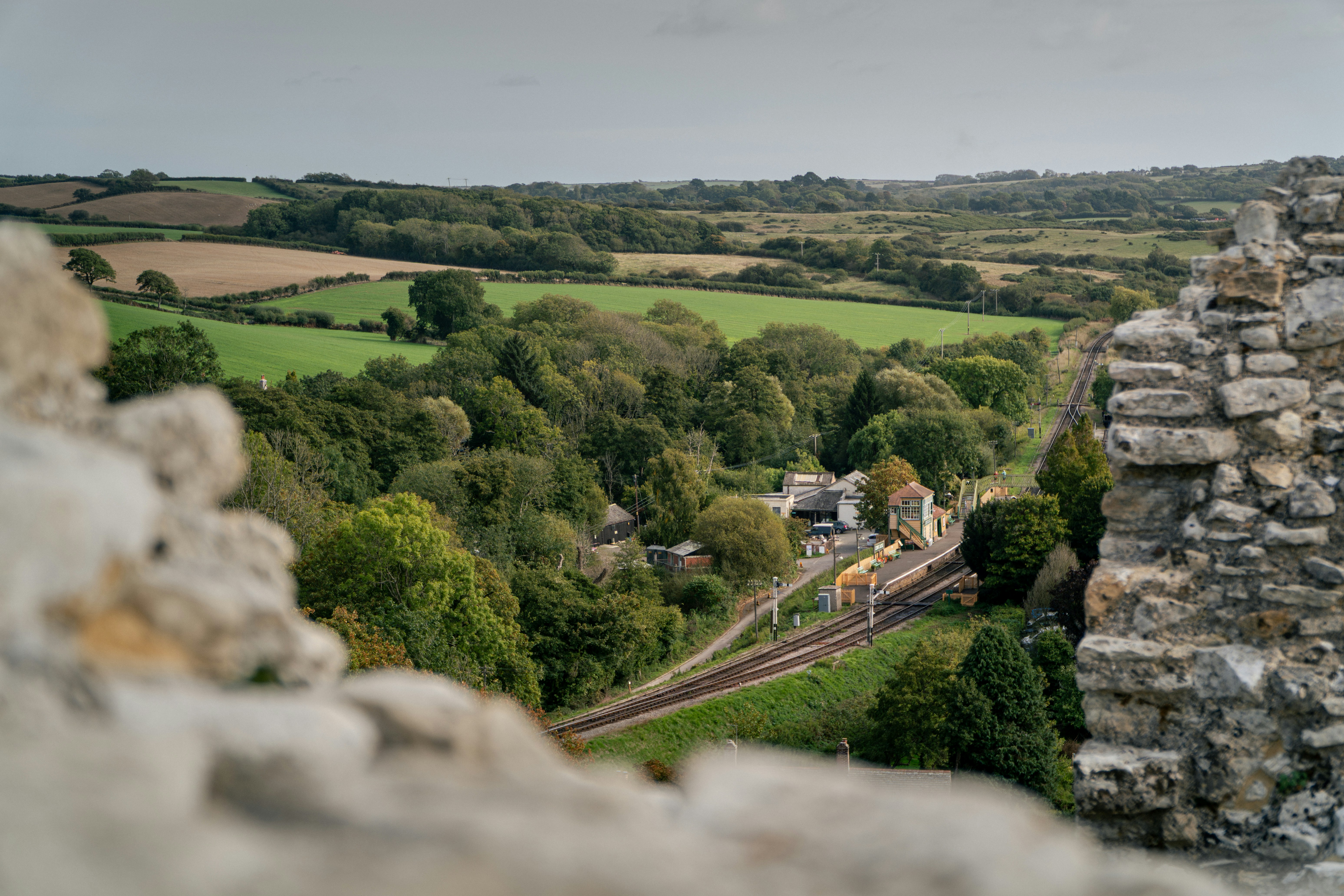 Small railway junction tucked among trees and fields, viewed from a crumbling stone vantage with distant village details.
