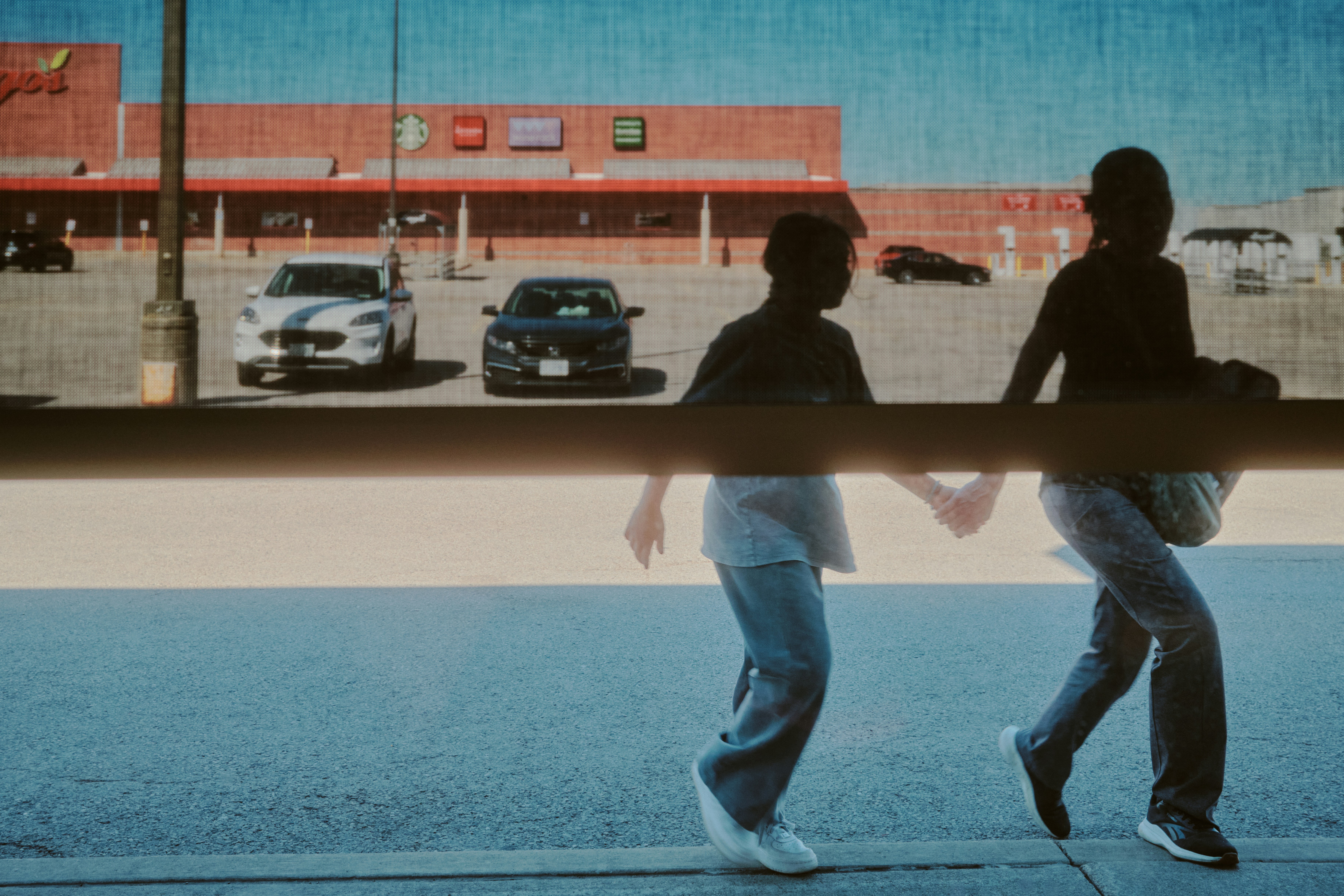 Two people walking hand in hand past a plaza parking lot on a summer afternoon.
