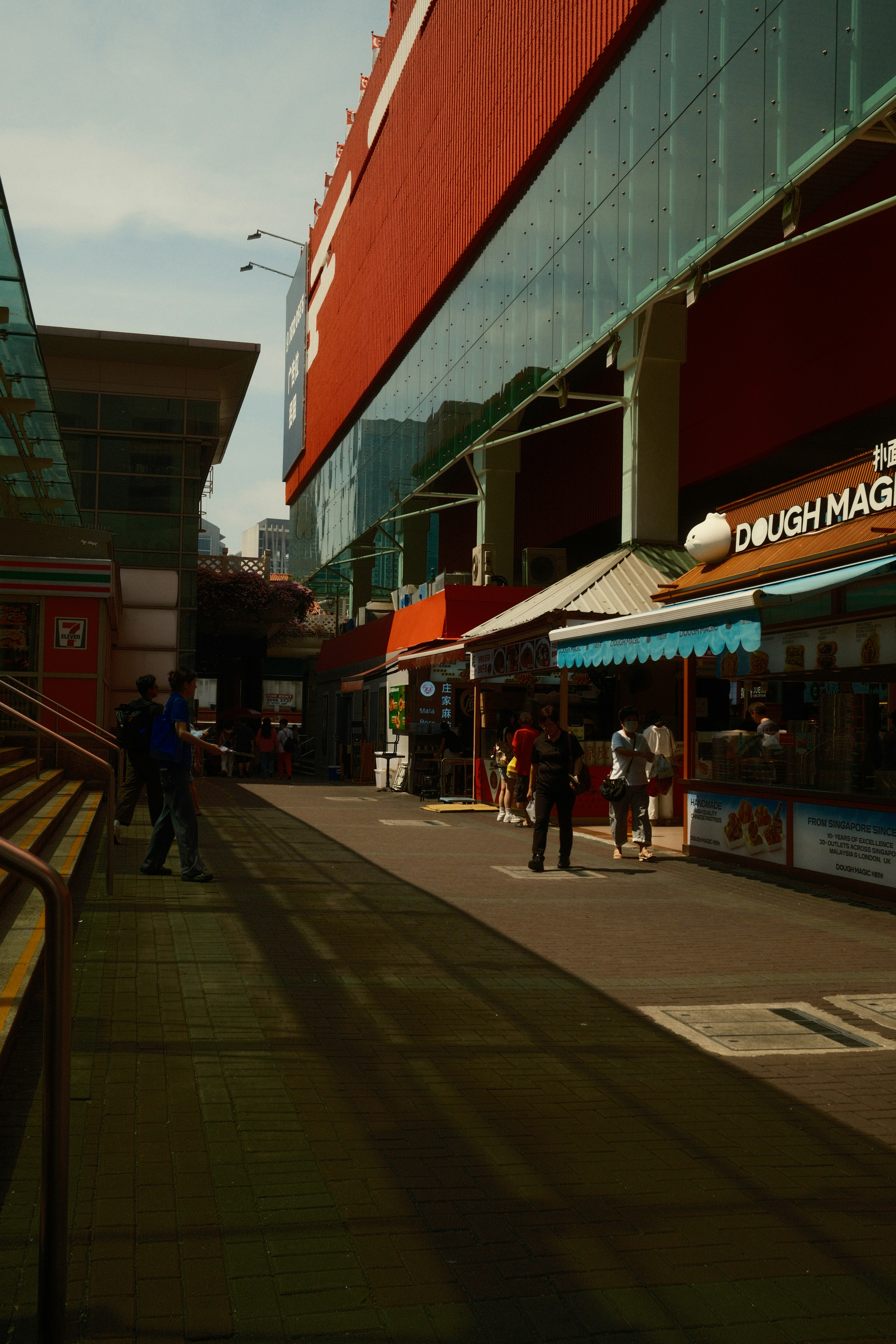 A sunlit urban walkway lined with storefronts and awnings, with a few pedestrians and shoppers moving along the street.