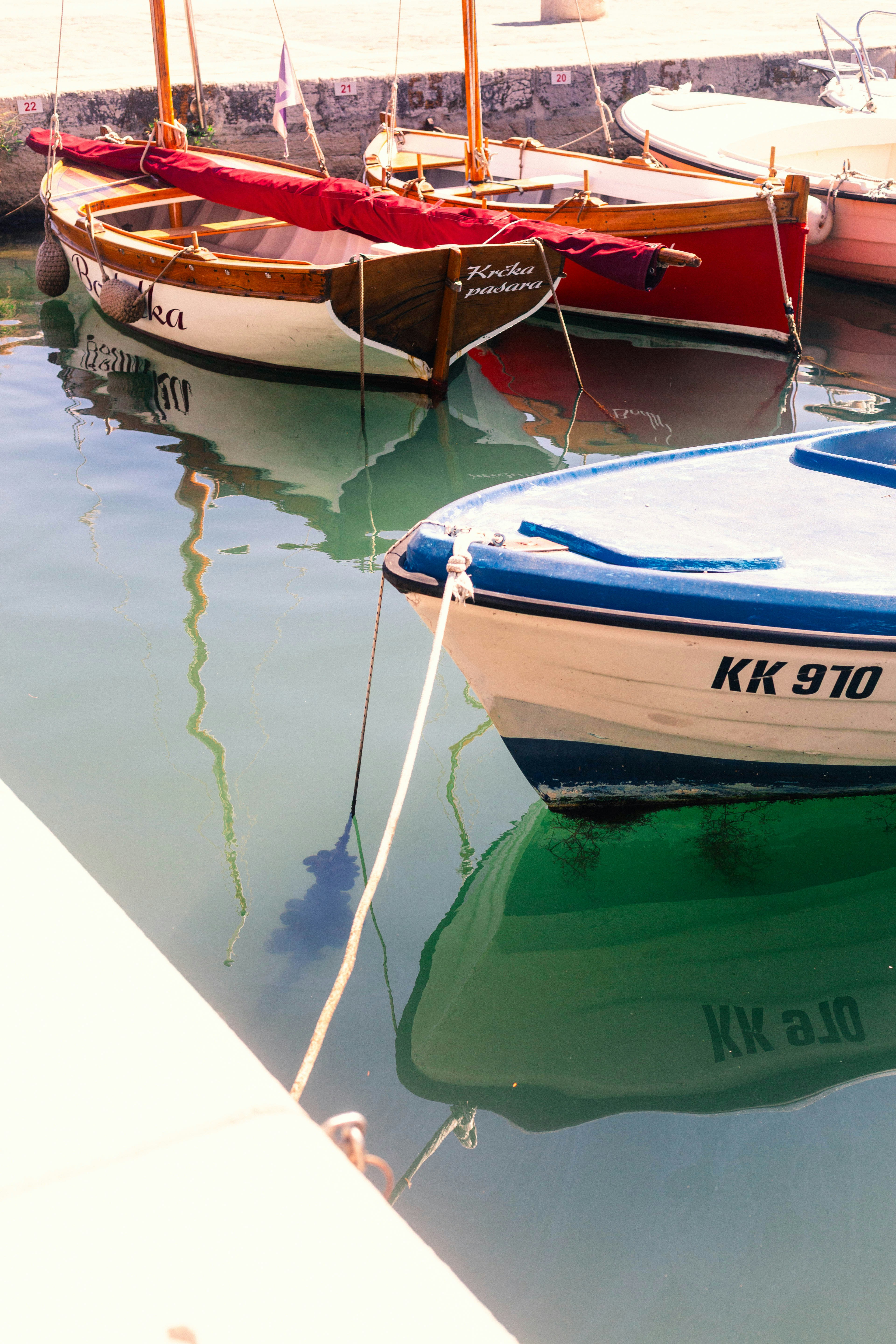Colorful boats gently bob in calm water, their reflections stretching across the glassy surface at a sunlit harbor.