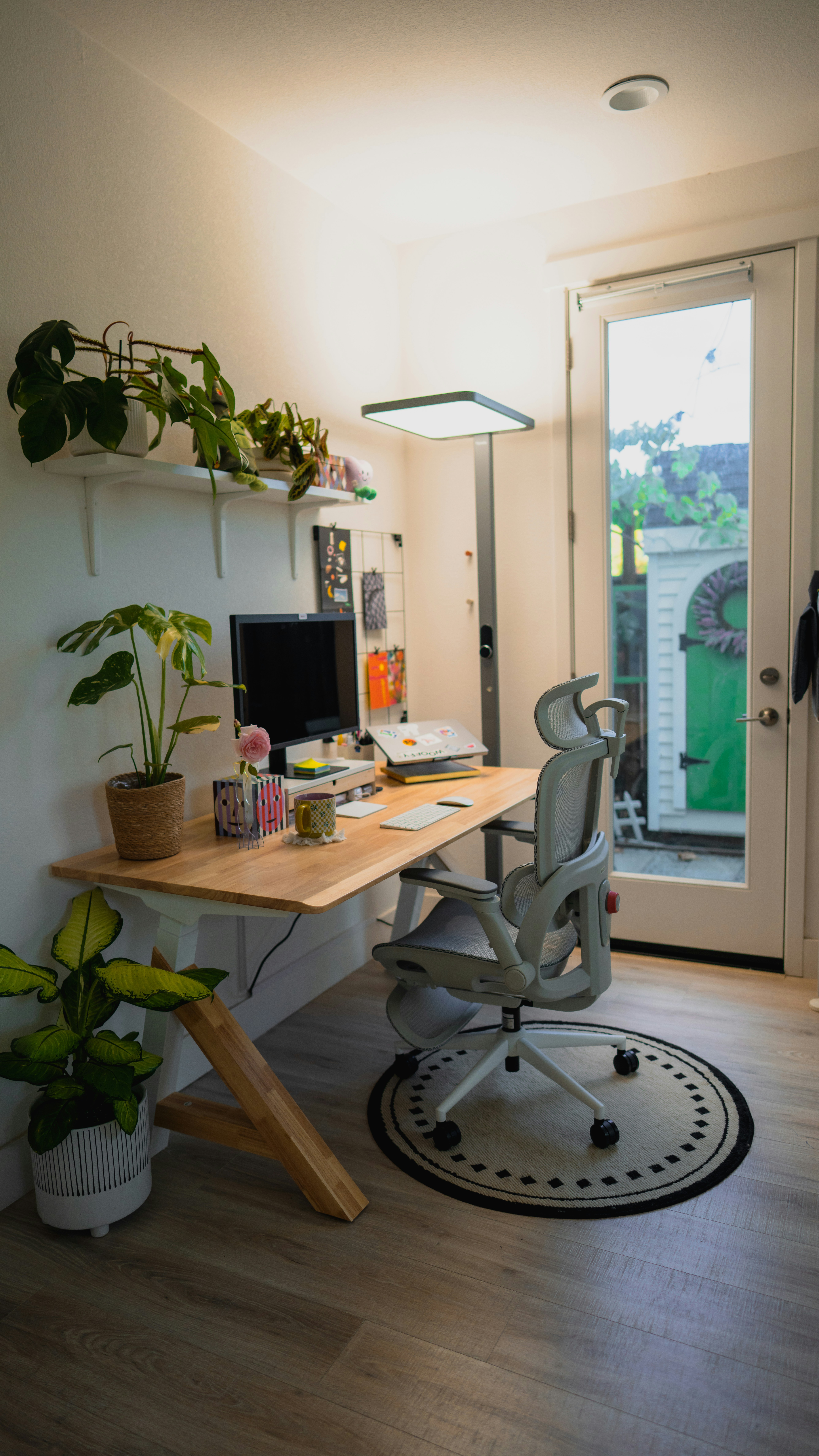 Bright home office with a wooden desk, ergonomic chair, and lush potted plants; large glass door opens to an outdoor area, bringing in natural light and a view of greenery. A rug, monitor, and organized shelves complete the workspace.