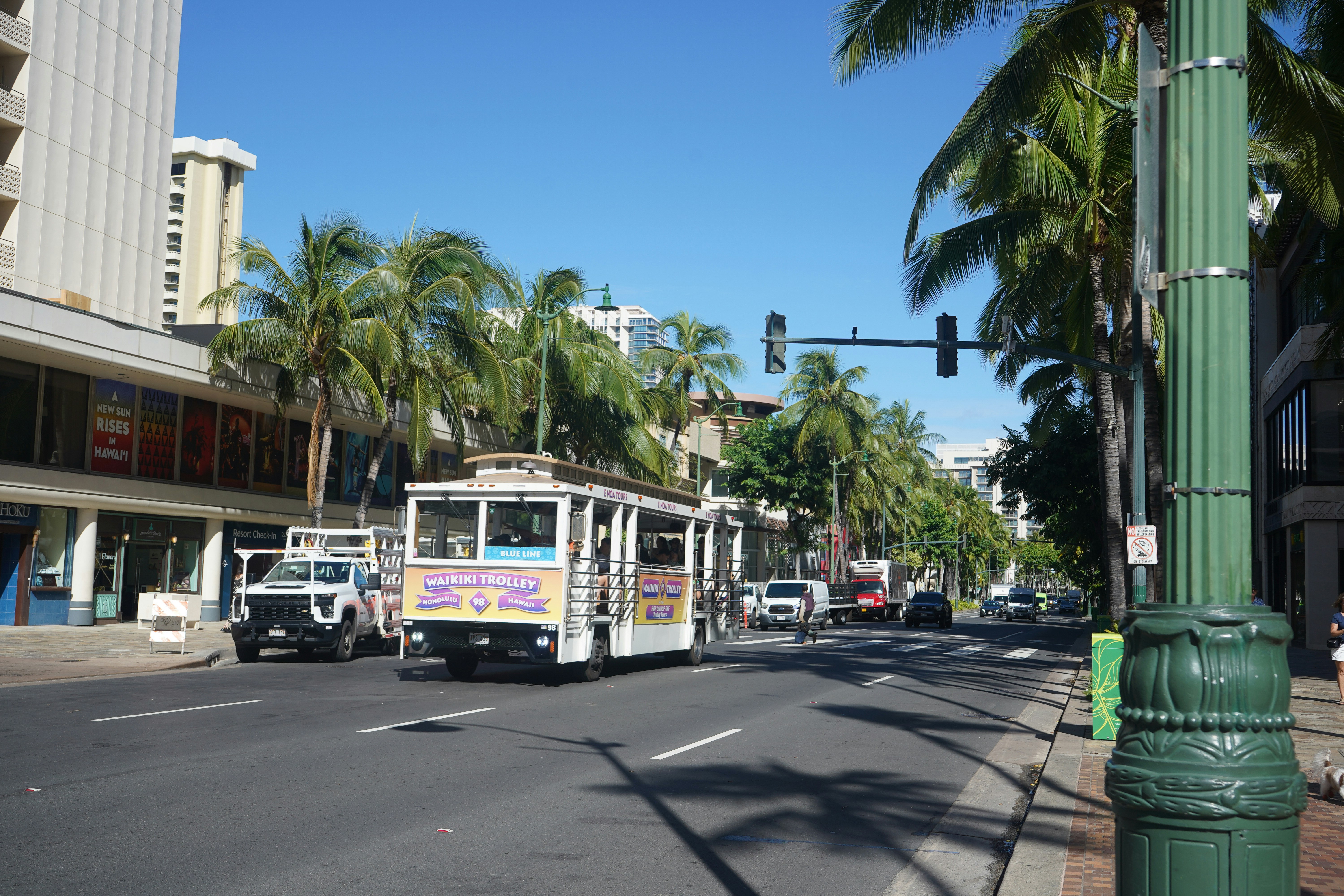 Sunlit city street with palm trees, a vintage trolley and traffic along a broad avenue.