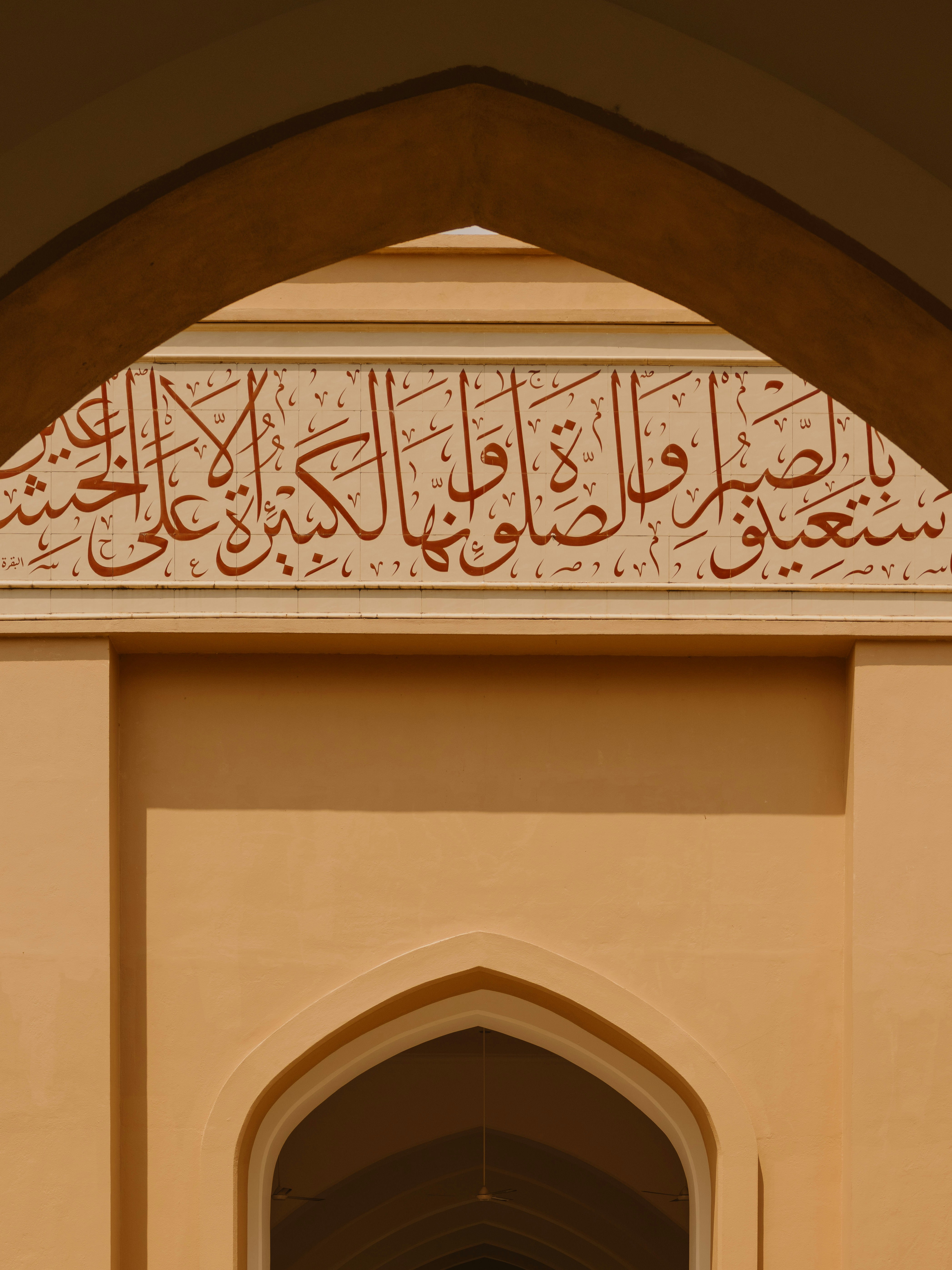 Arabic calligraphy carved above a pointed arch at Masjid Tengku Ampuan in Kuala Lumpur, with soft warm light on the sandstone walls.