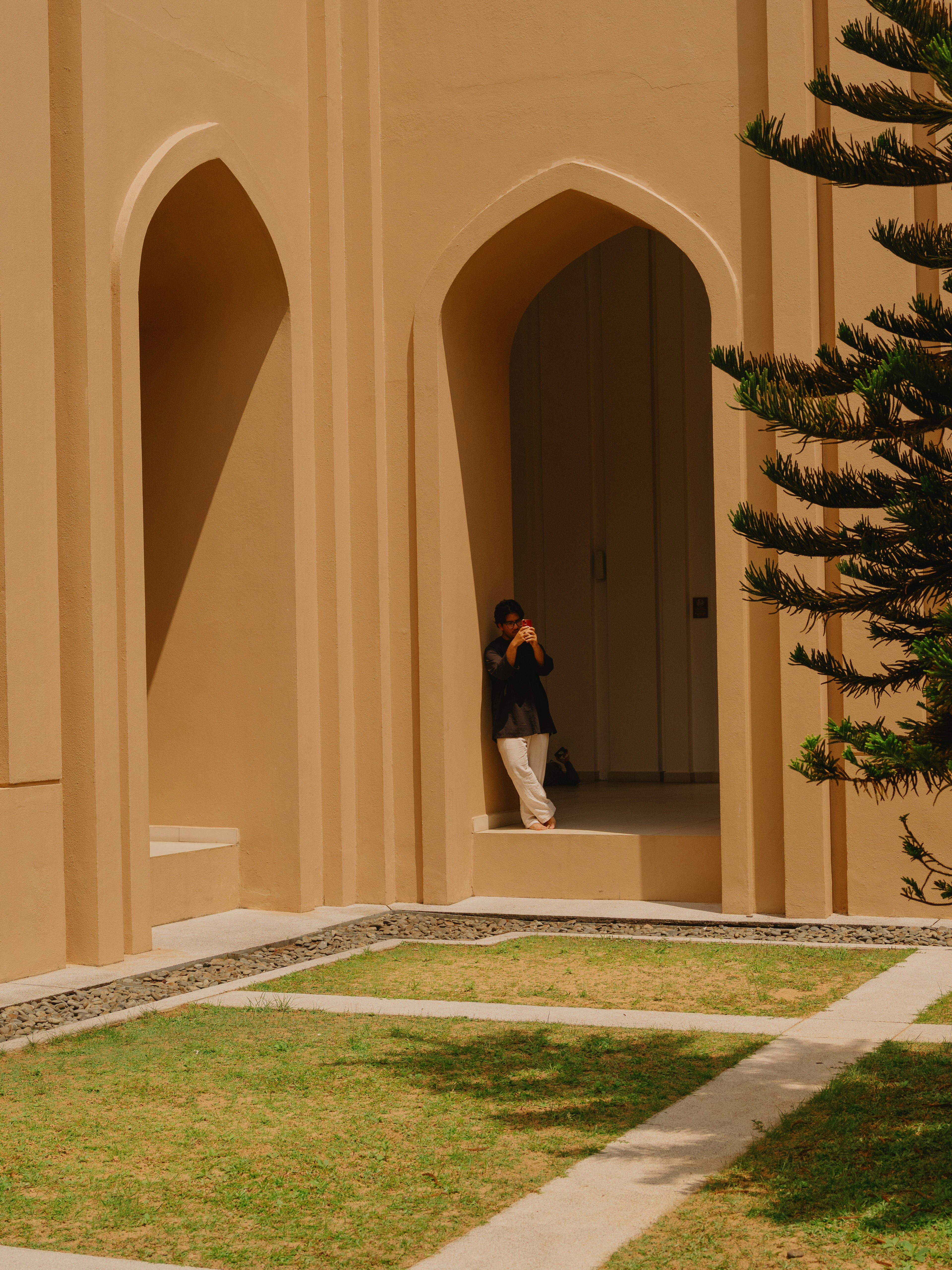 A quiet courtyard at Masjid Tengku Ampuan in Kuala Lumpur, where a photographer frames a shot beneath tall arches beside a green lawn and pine tree.