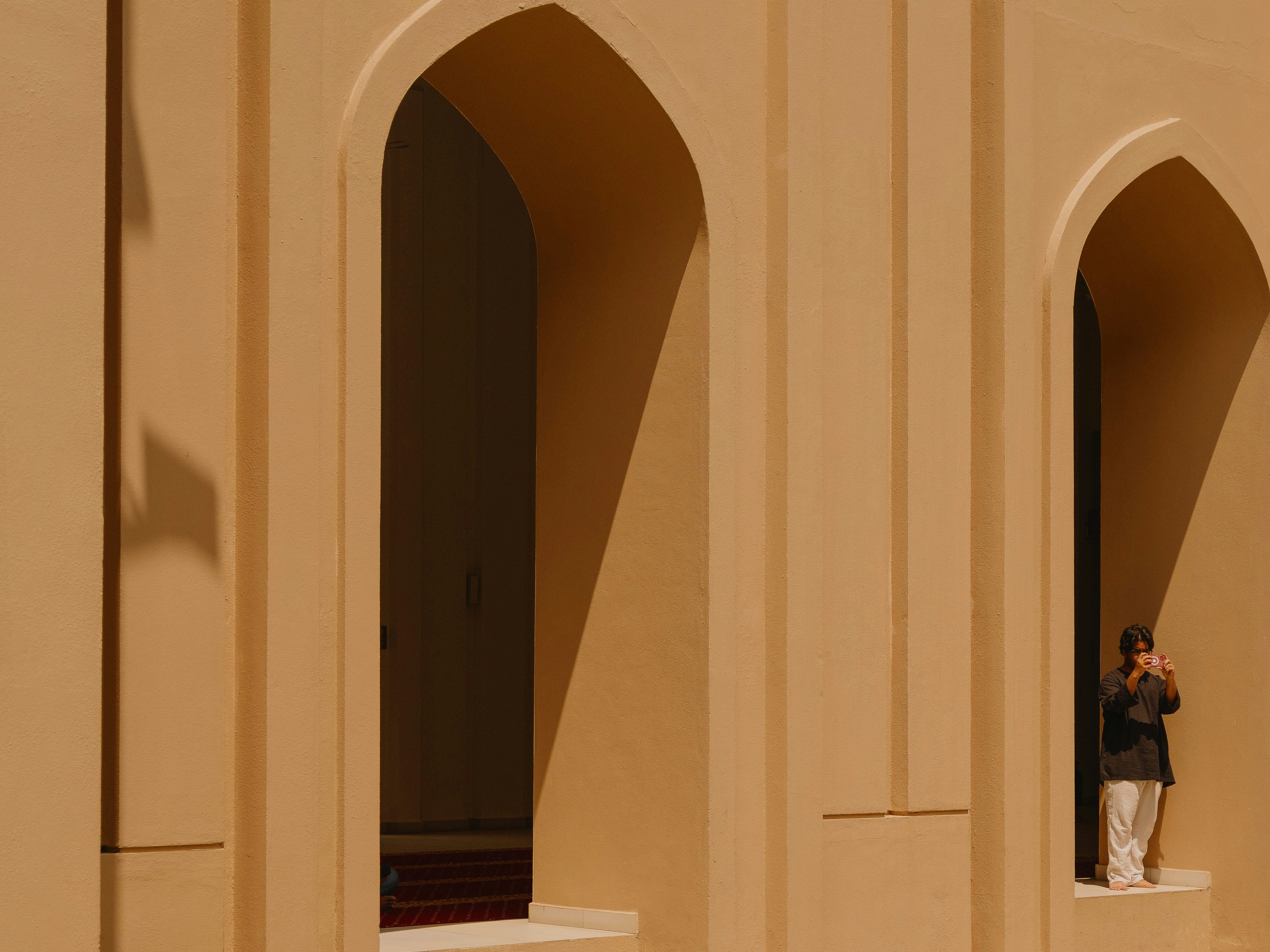 A photographer stands in the shade of a tall pointed doorway at Masjid Tengku Ampuan, dwarfed by the warm sandstone walls.