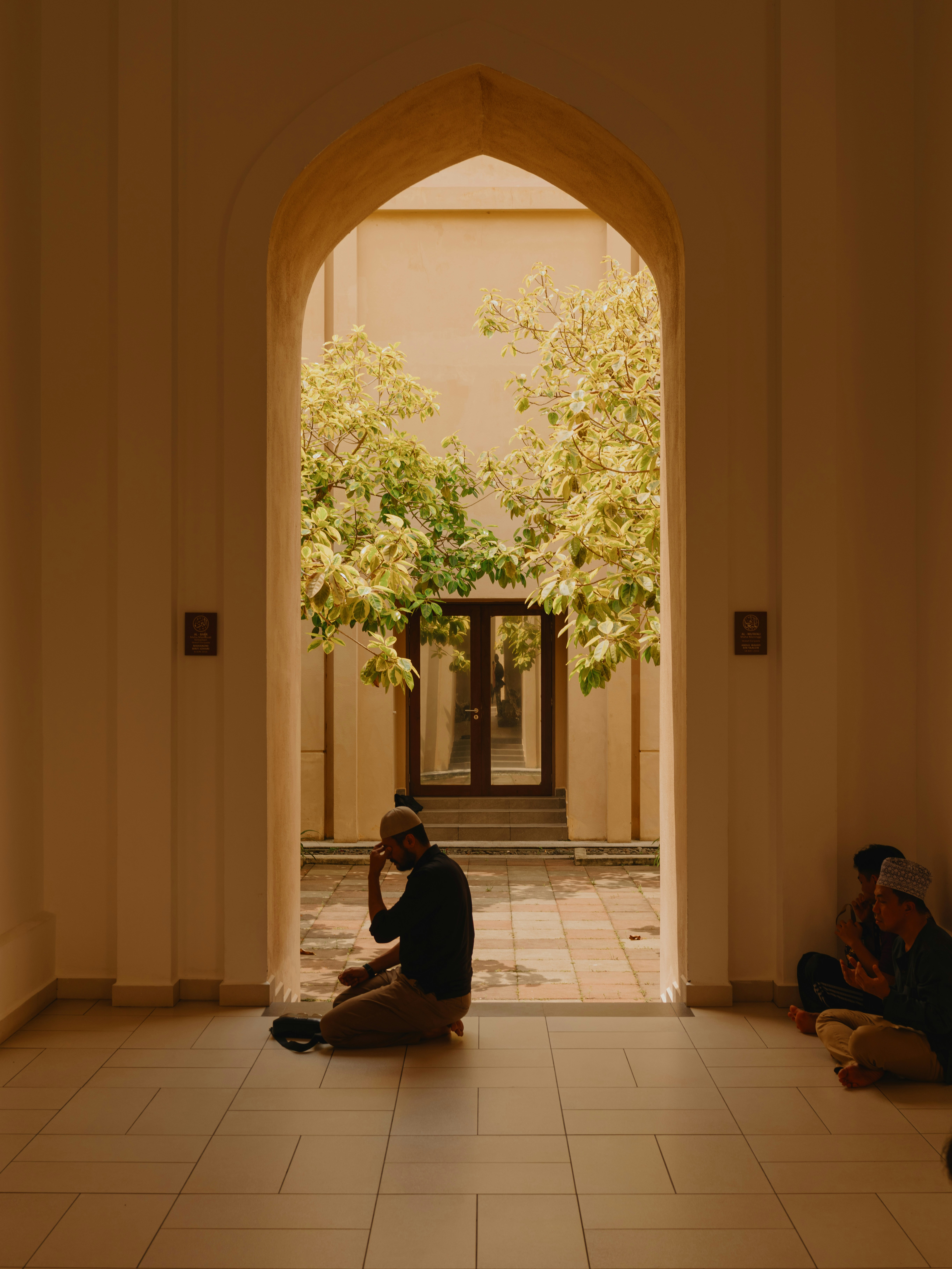 A man sits in quiet prayer along a corridor at Masjid Tengku Ampuan, with a pointed arch opening onto a leafy courtyard in soft afternoon light.