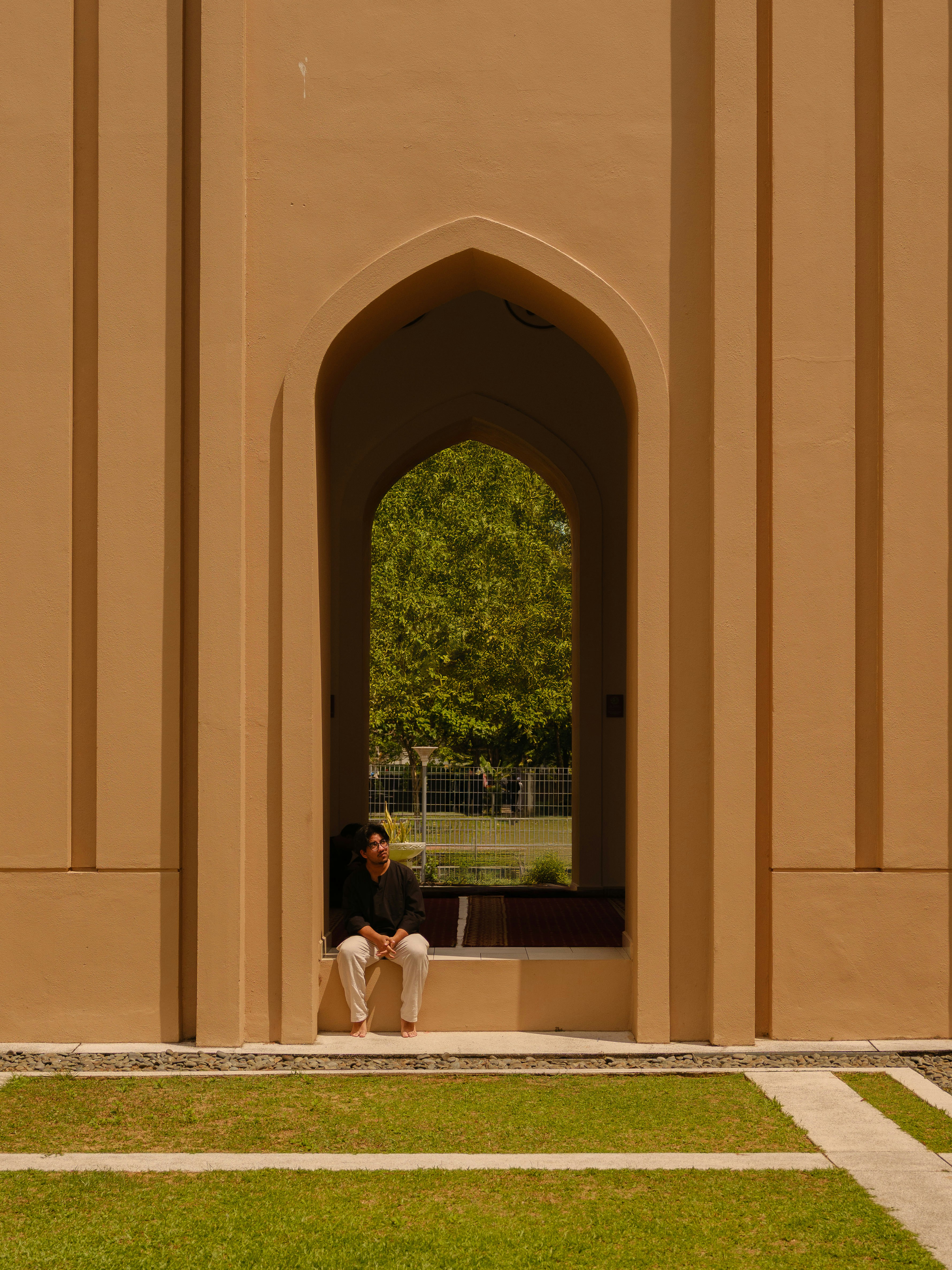 A man sits quietly beneath a pointed archway at Masjid Tengku Ampuan, with trees and green lawn framed by the warm sandstone walls.
