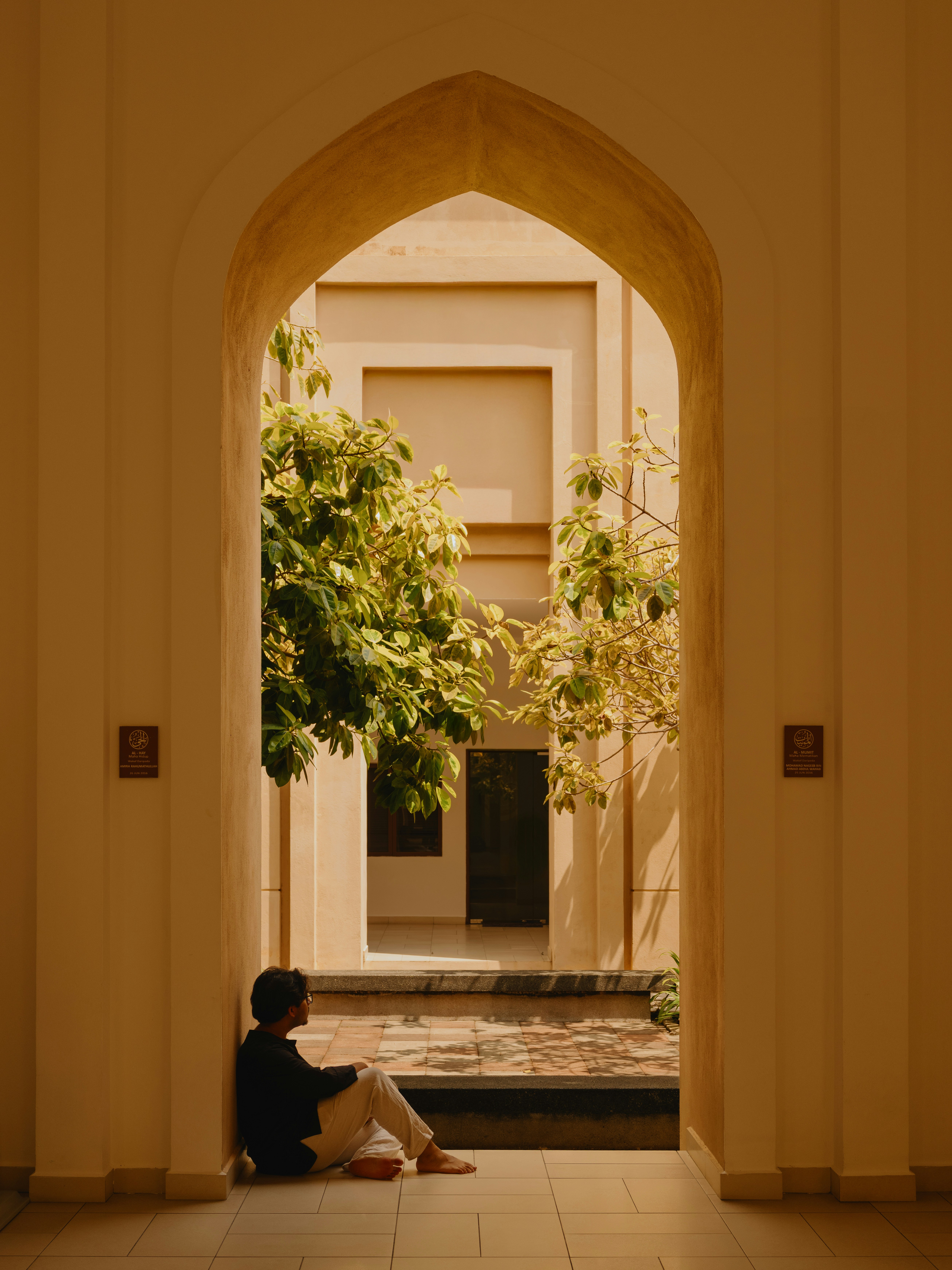 A person rests beneath a pointed archway at Masjid Tengku Ampuan, looking out onto a leafy courtyard bathed in warm light.