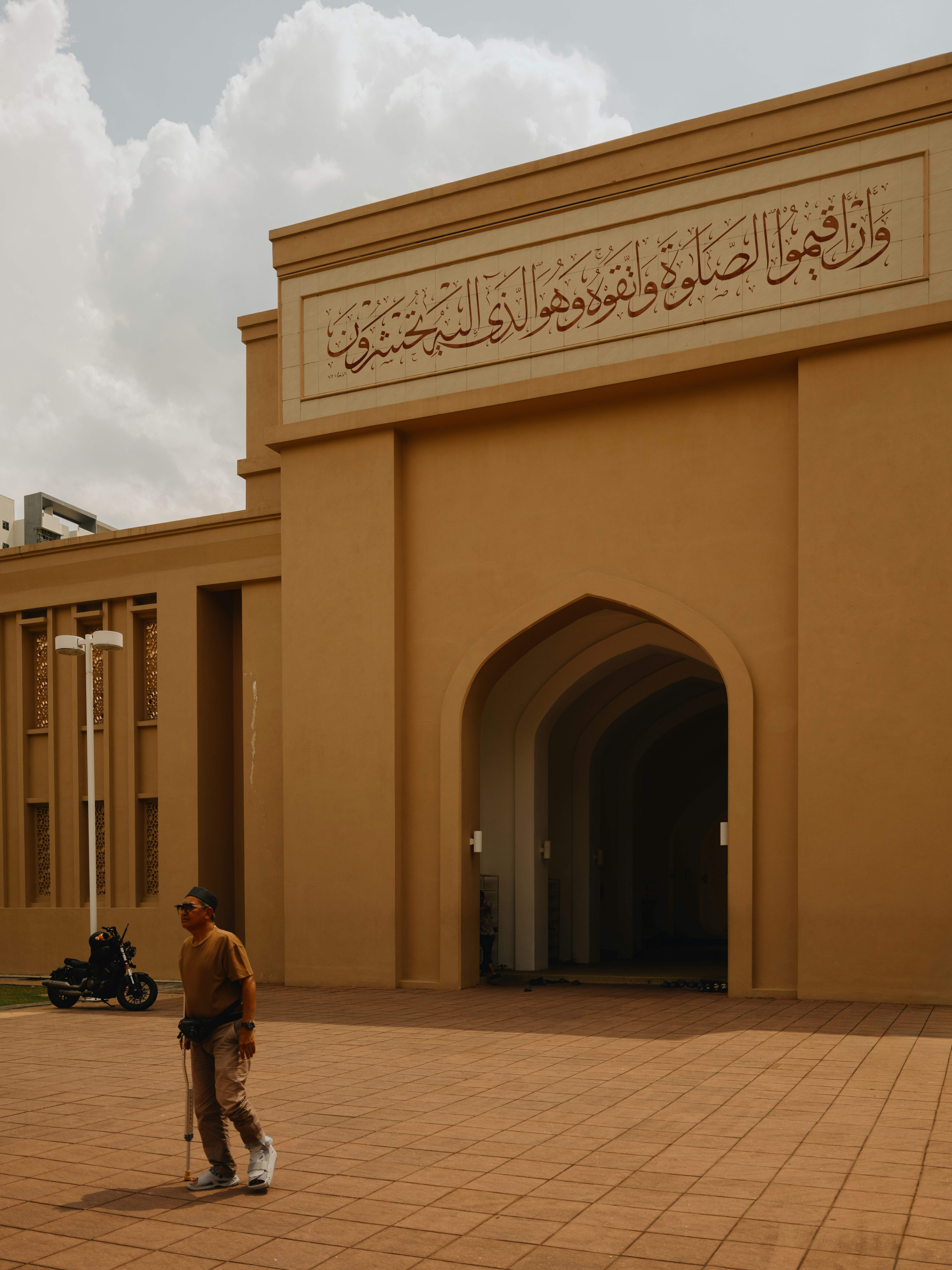 The calligraphy lined entrance gate of Masjid Tengku Ampuan in Kuala Lumpur, with a man strolling past under a cloudy afternoon sky.