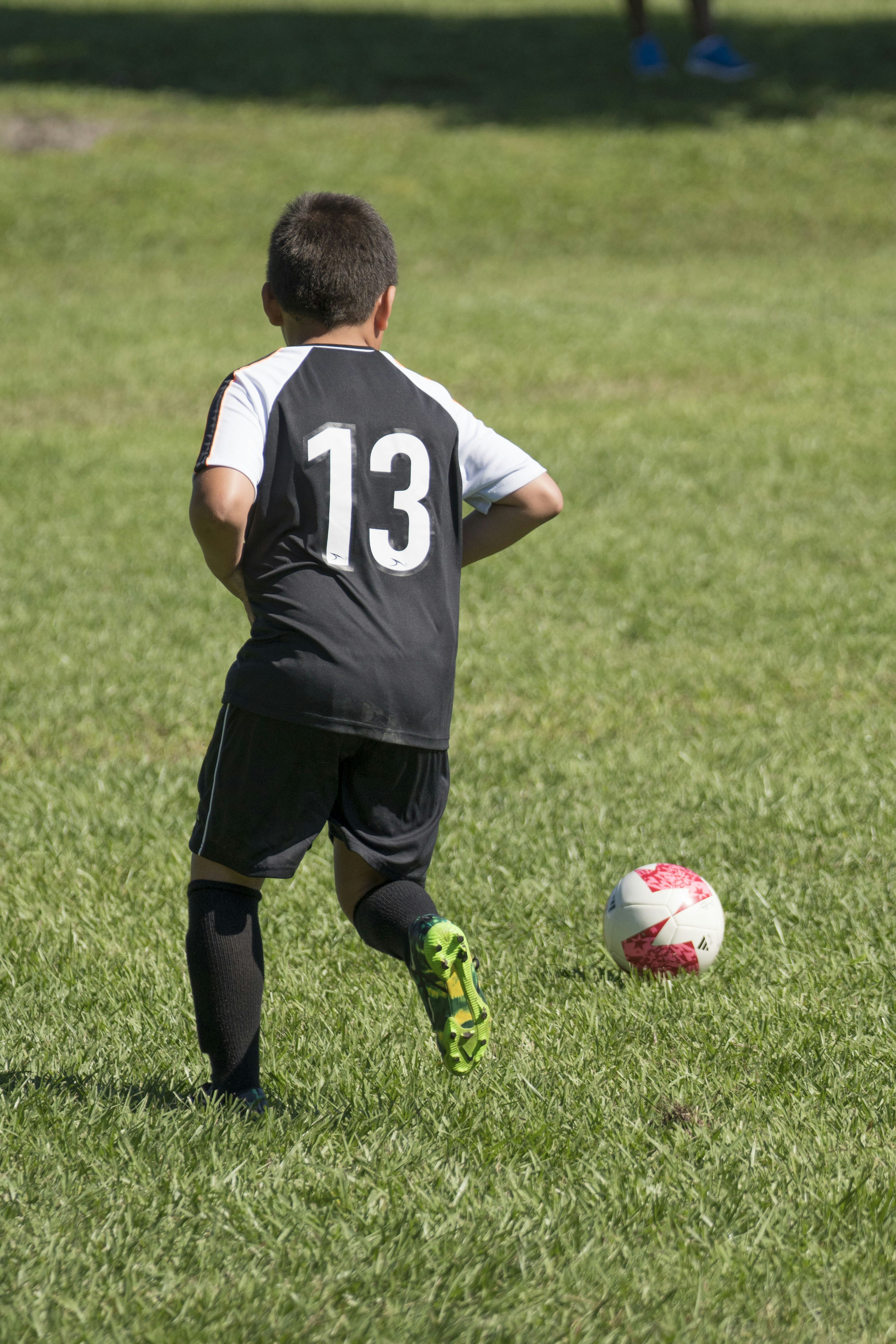 Soccer player in a black jersey with the number 13 dribbles toward a ball on a sunlit field; empty green grass dominates the background and partial legs of another player appear at the top.