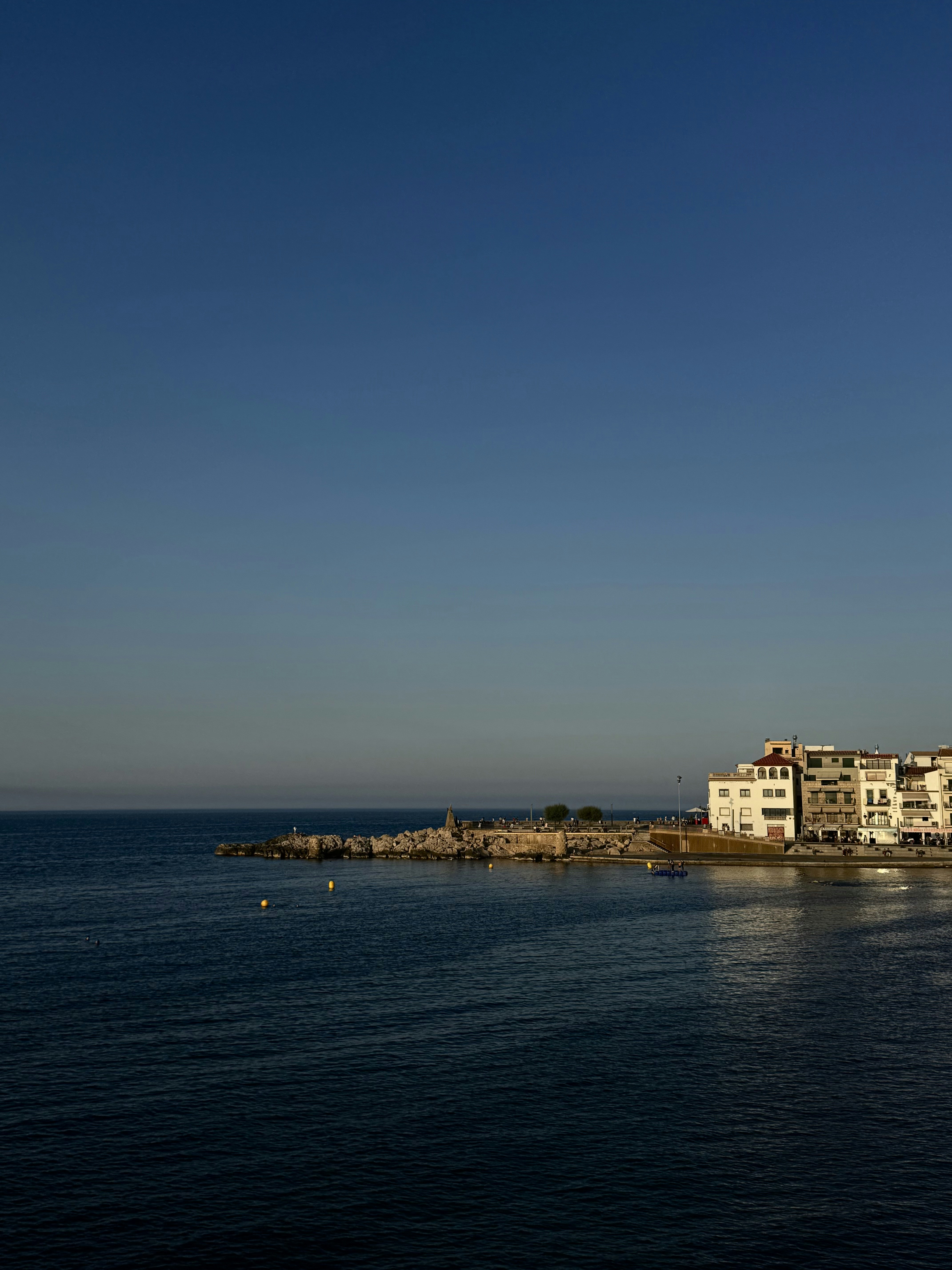 Harbor shoreline with calm water and a small cluster of buildings along the quay, under a clear gradient sky; the scene emphasizes the coastal town edge and water reflections.