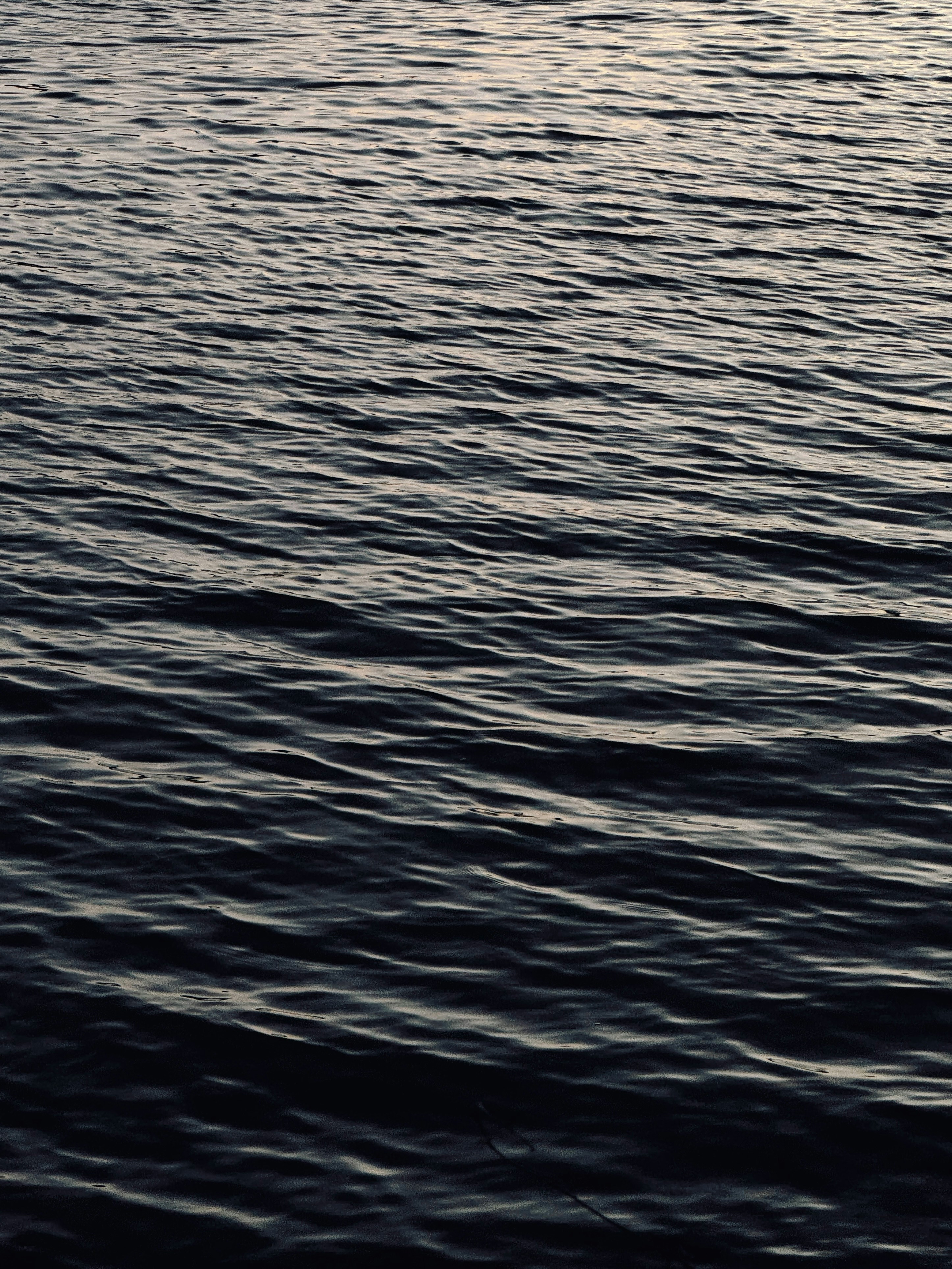 Close-up of dark, gently undulating water with subtle highlights from fading light; a minimal, abstract capture of waves and texture. This is a water surface photograph, likely photographed from above or at a shallow angle.