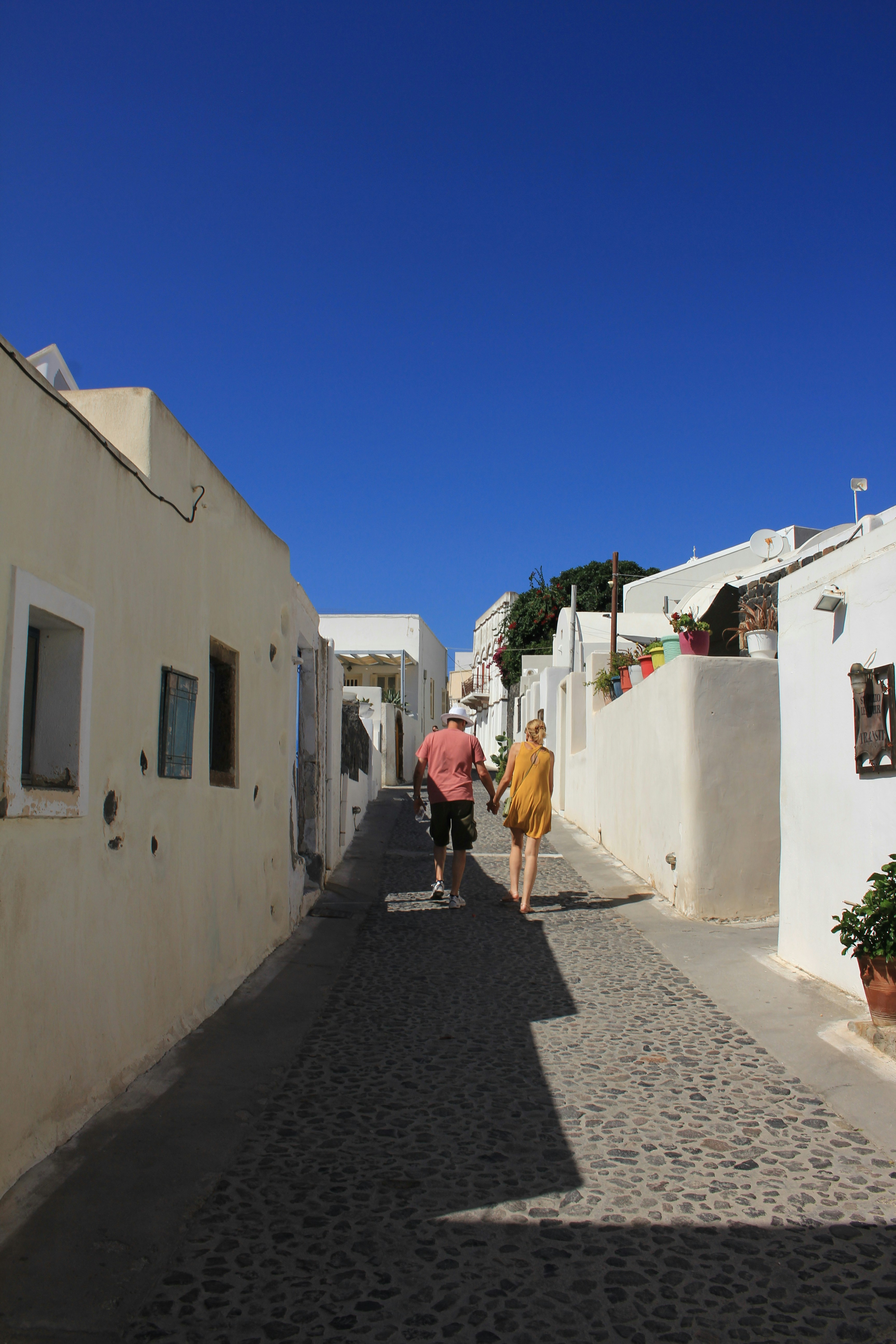 Two people walk arm-in-arm down a sunlit cobblestone alley flanked by white-washed buildings under a clear blue sky.