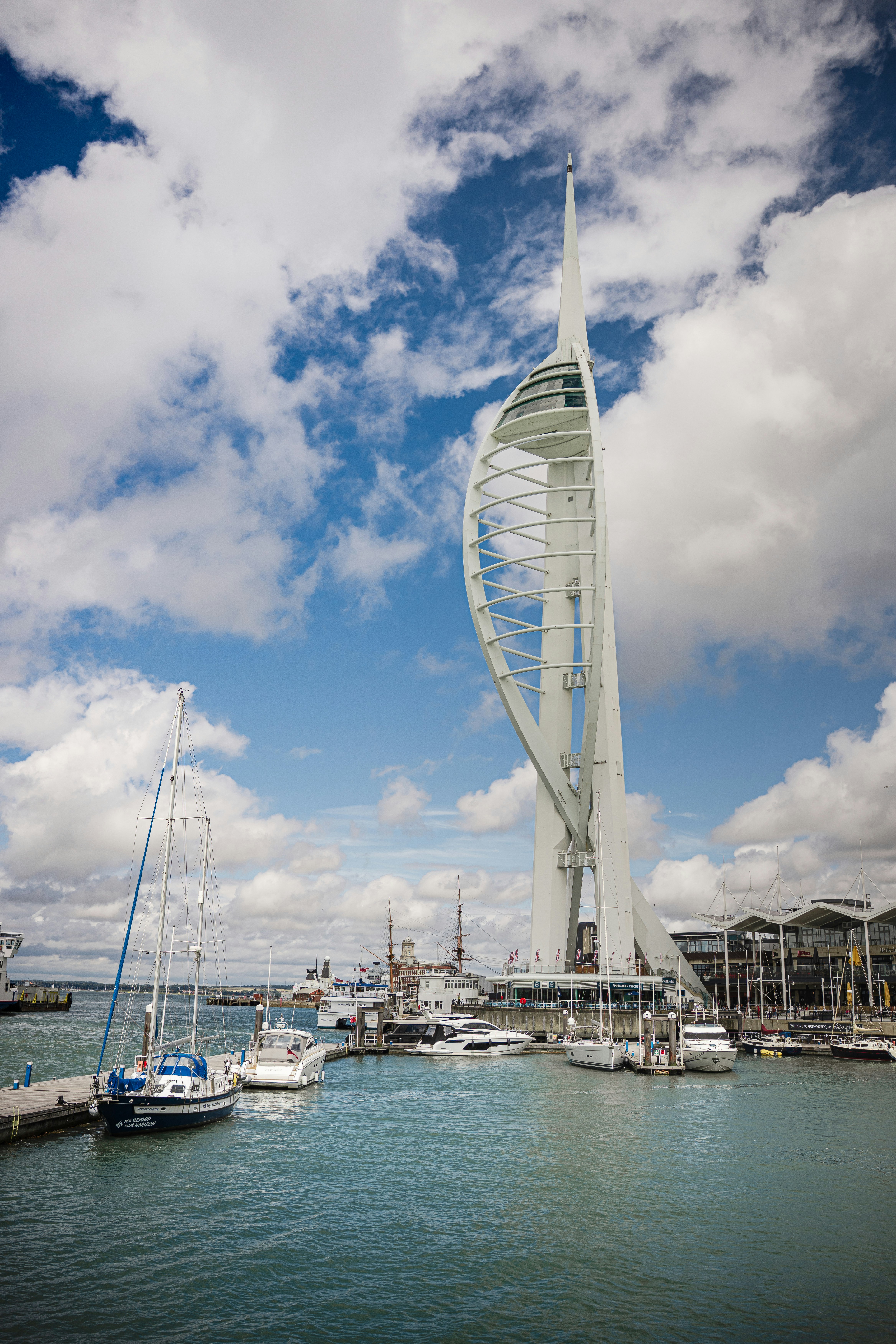 Marina at a sunny harbor with moored boats and a tall sail-shaped tower rising over the docks at modest distance.