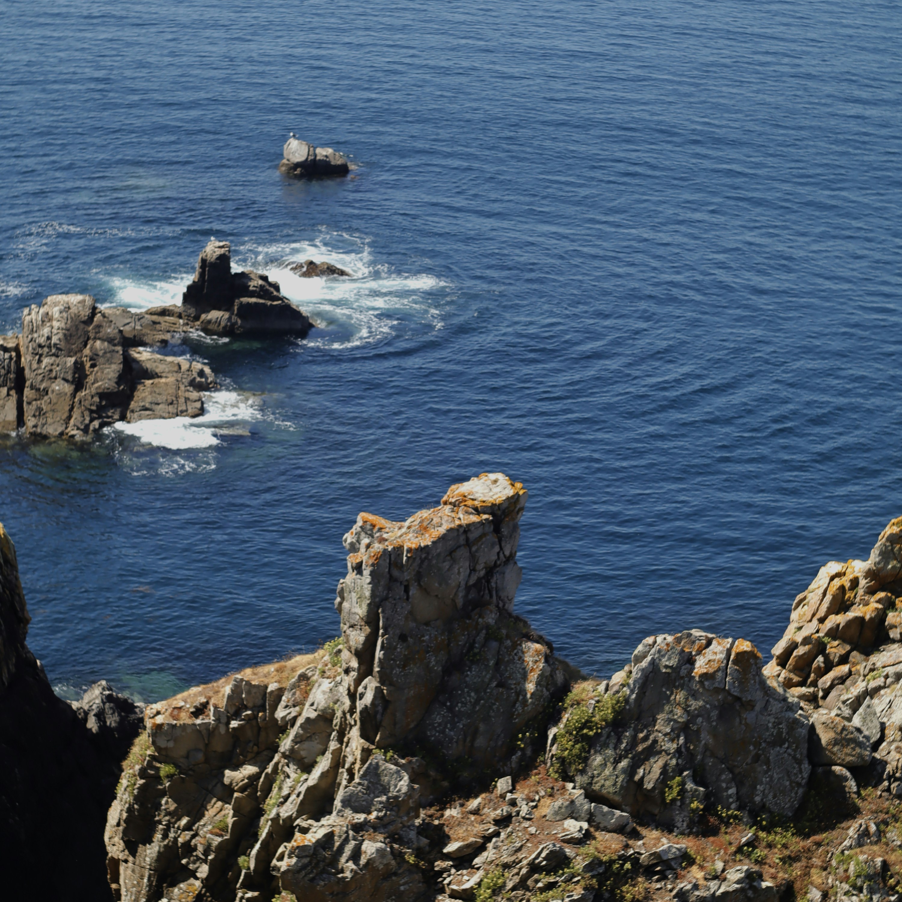 Sunlit jagged rocks meet deep blue sea along a rugged coastline, with waves brushing the base of the formations. A natural seascape emphasizing texture and color without people or wildlife.