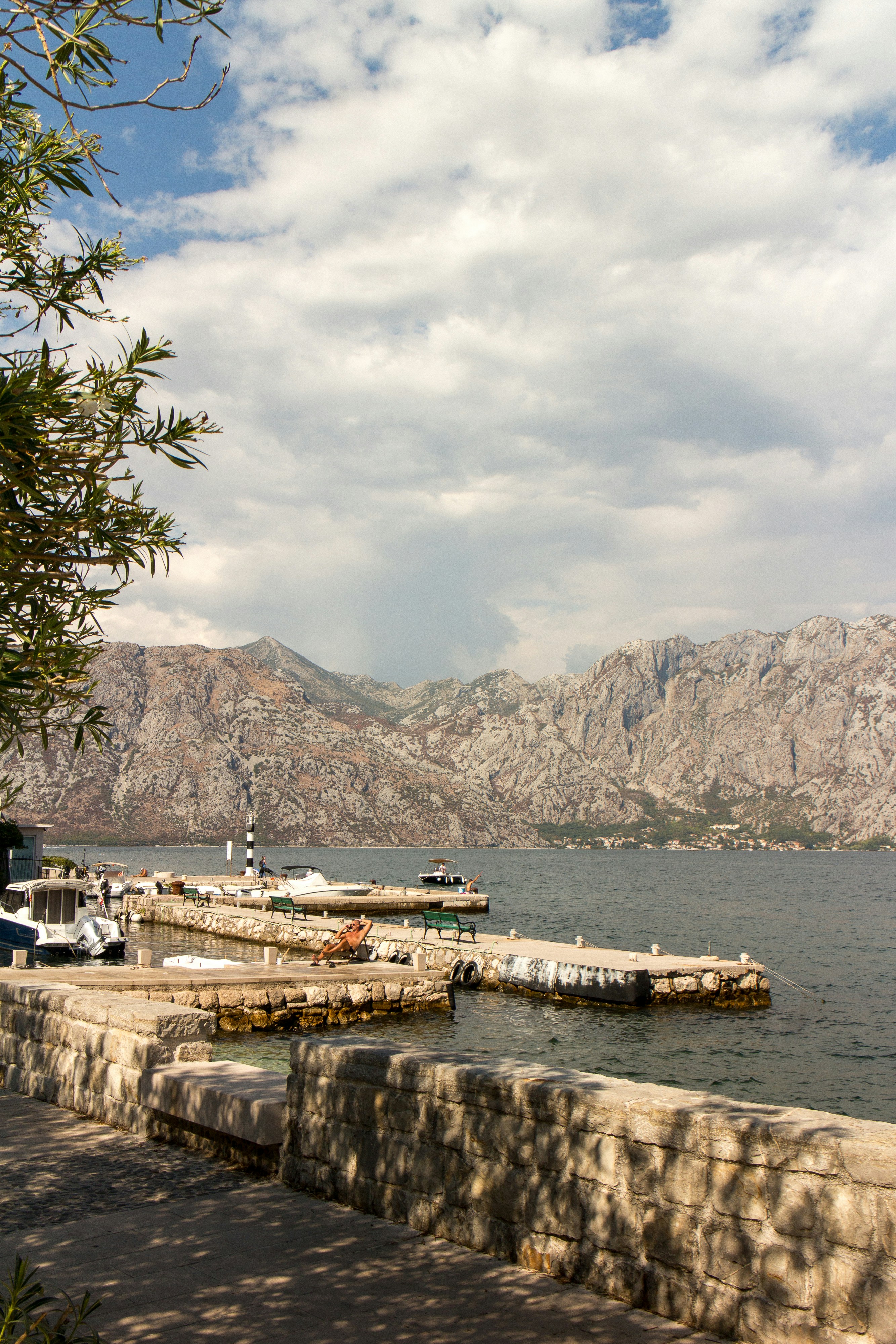 Harbor scene with a stone jetty and moored boats set against rugged rocky mountains under a partly cloudy sky.