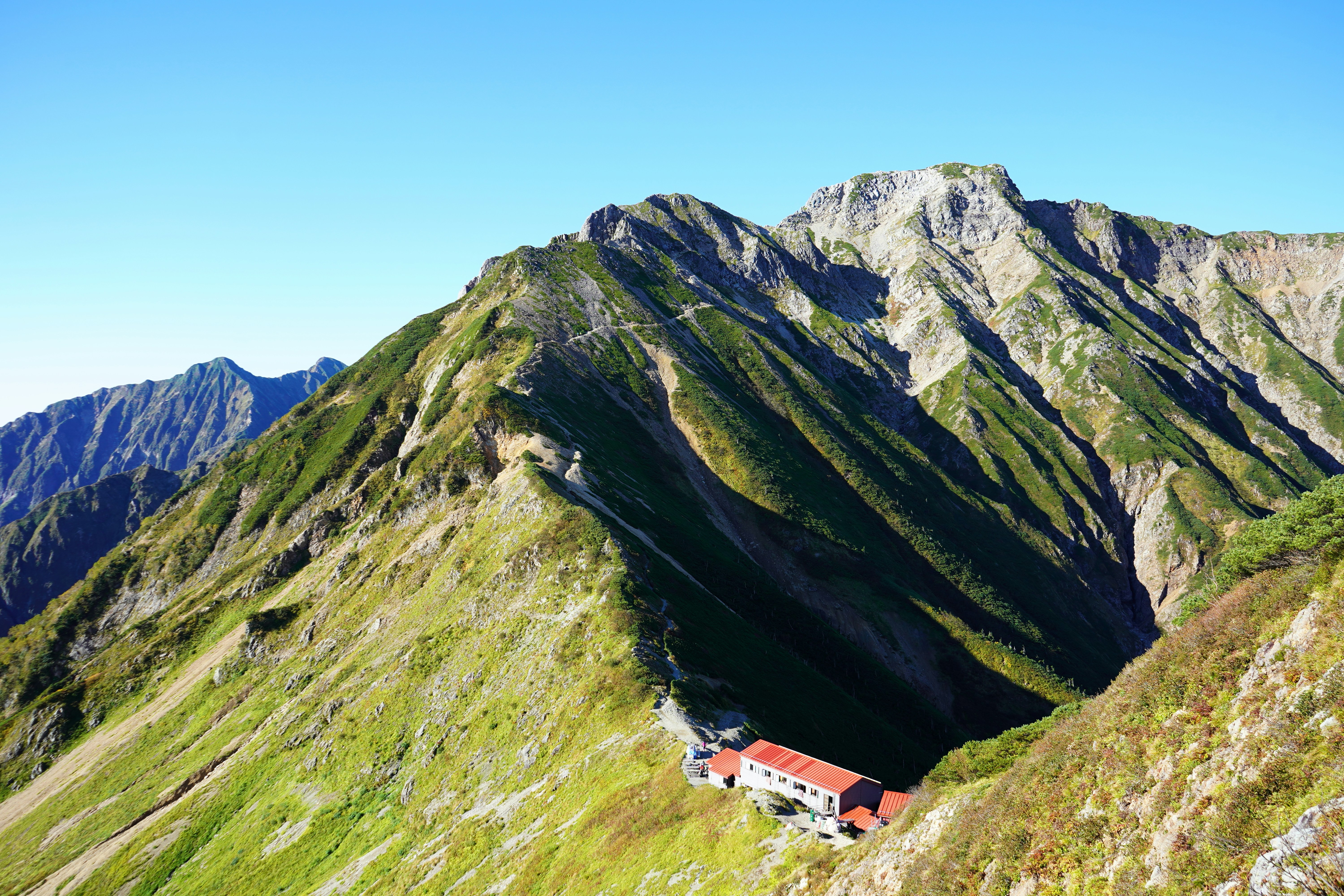 Bright ridge-top scene with a lone red-roofed building tucked into a grassy slope beneath jagged peaks.