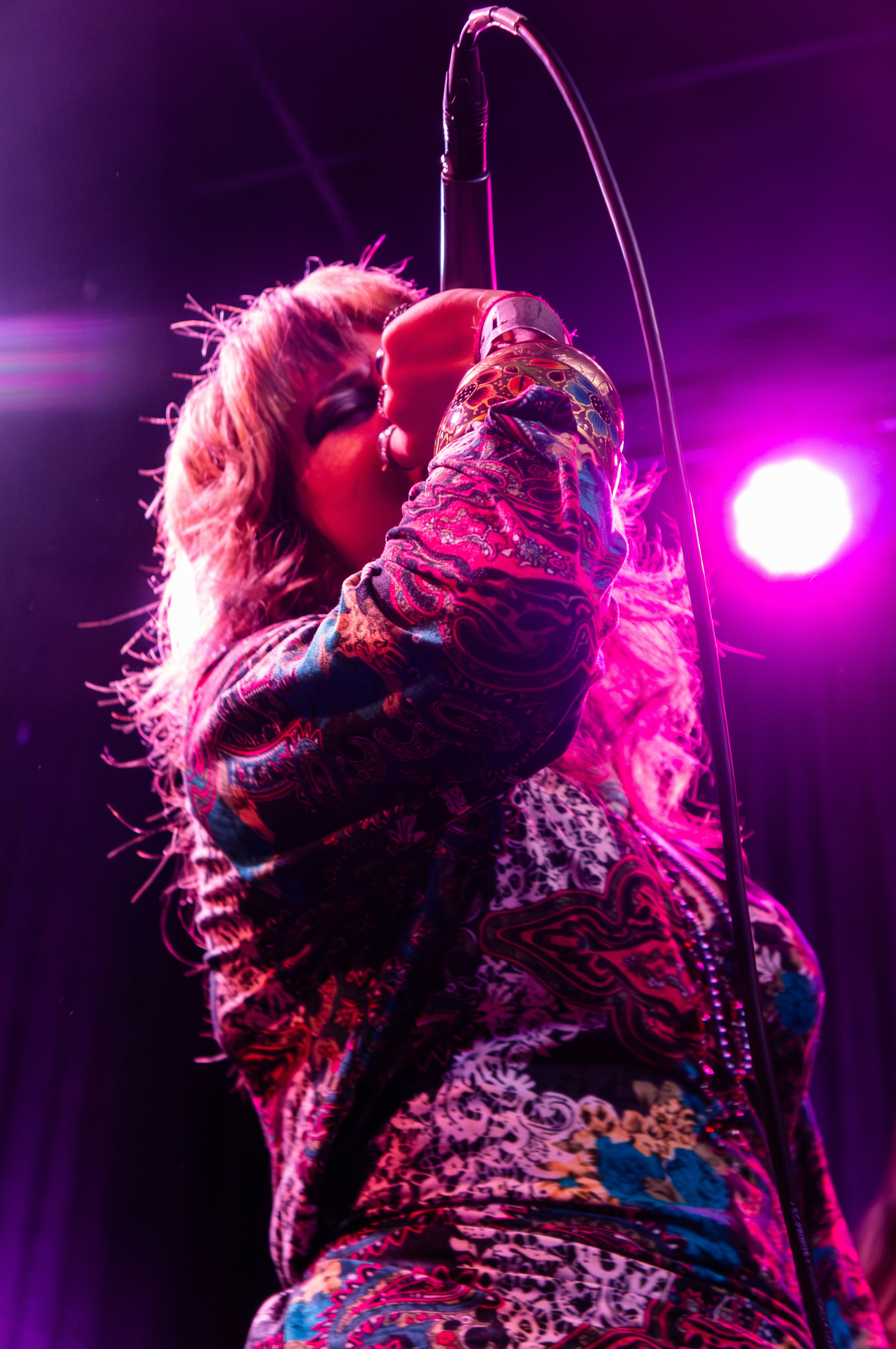 Energetic vocalist under magenta stage lighting, microphone close-up with intricate patterned attire and dynamic pose.