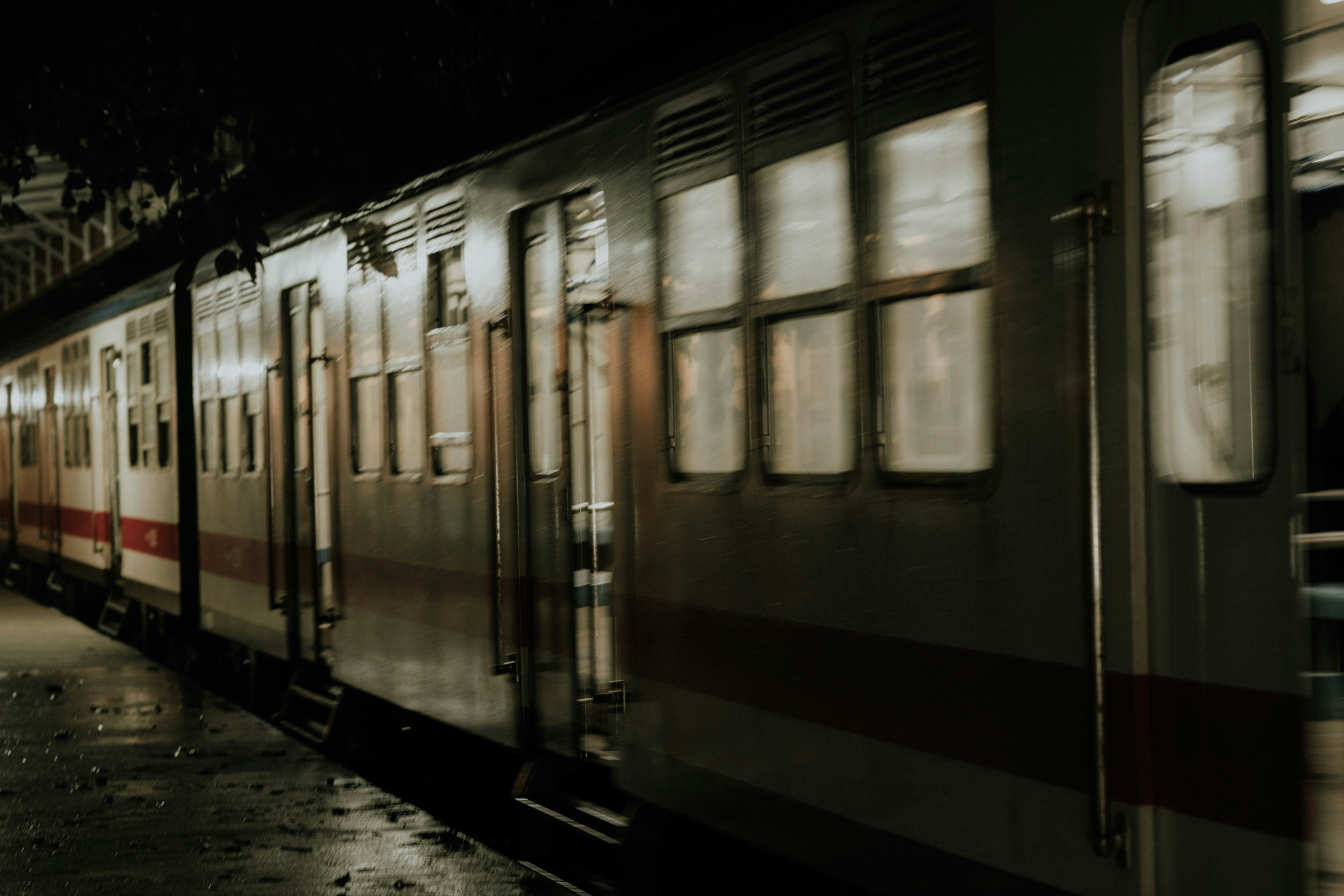 Night-time train carriages line a dark platform with dim light seeping through the windows. A solitary, muted scene focuses on the length of the train and the reflective wet ground.
