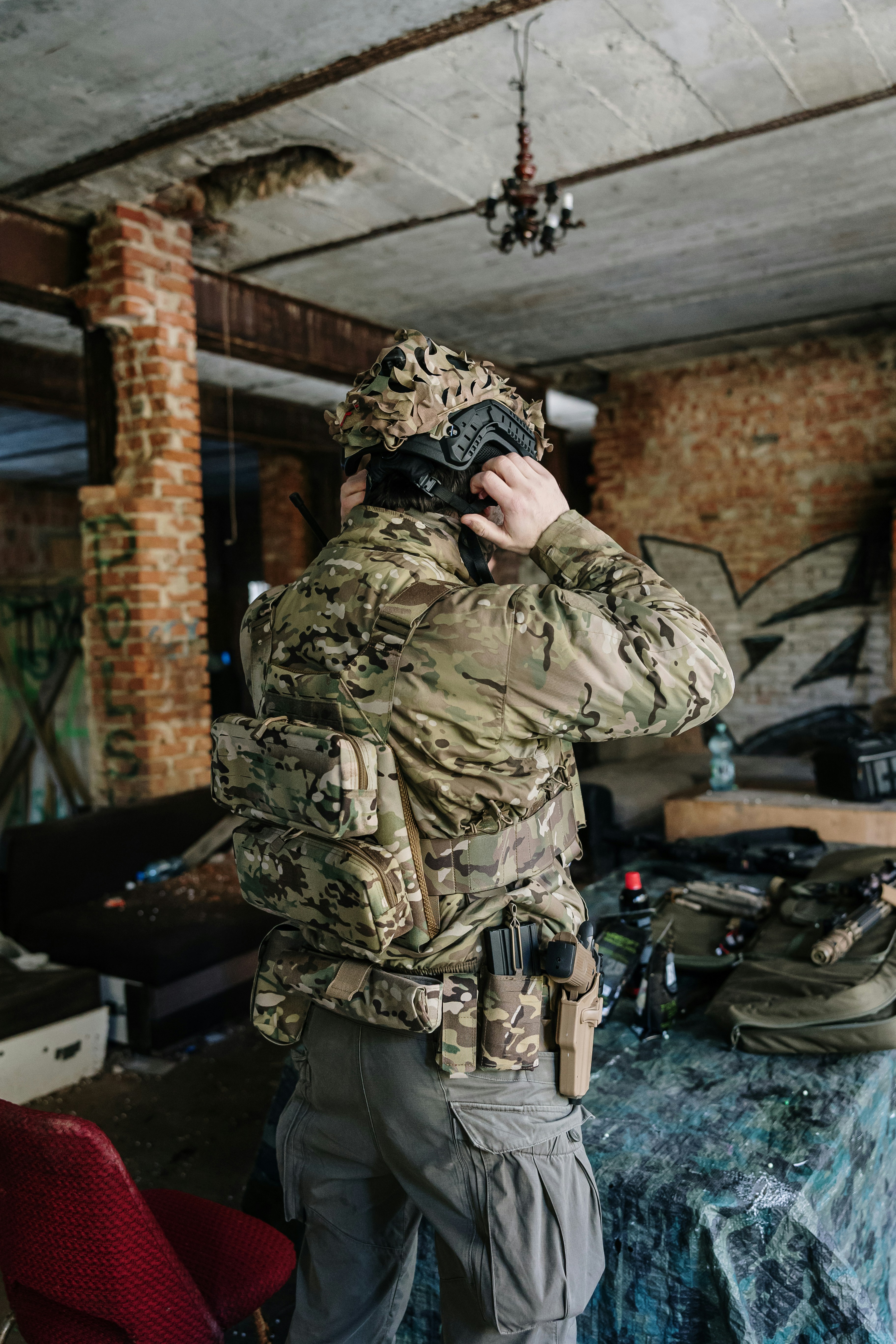 Camo-clad operator straps on gear inside an abandoned, graffiti-adorned space with brick pillars and scattered equipment. The scene emphasizes preparation and utilitarian detail in a gritty environment.