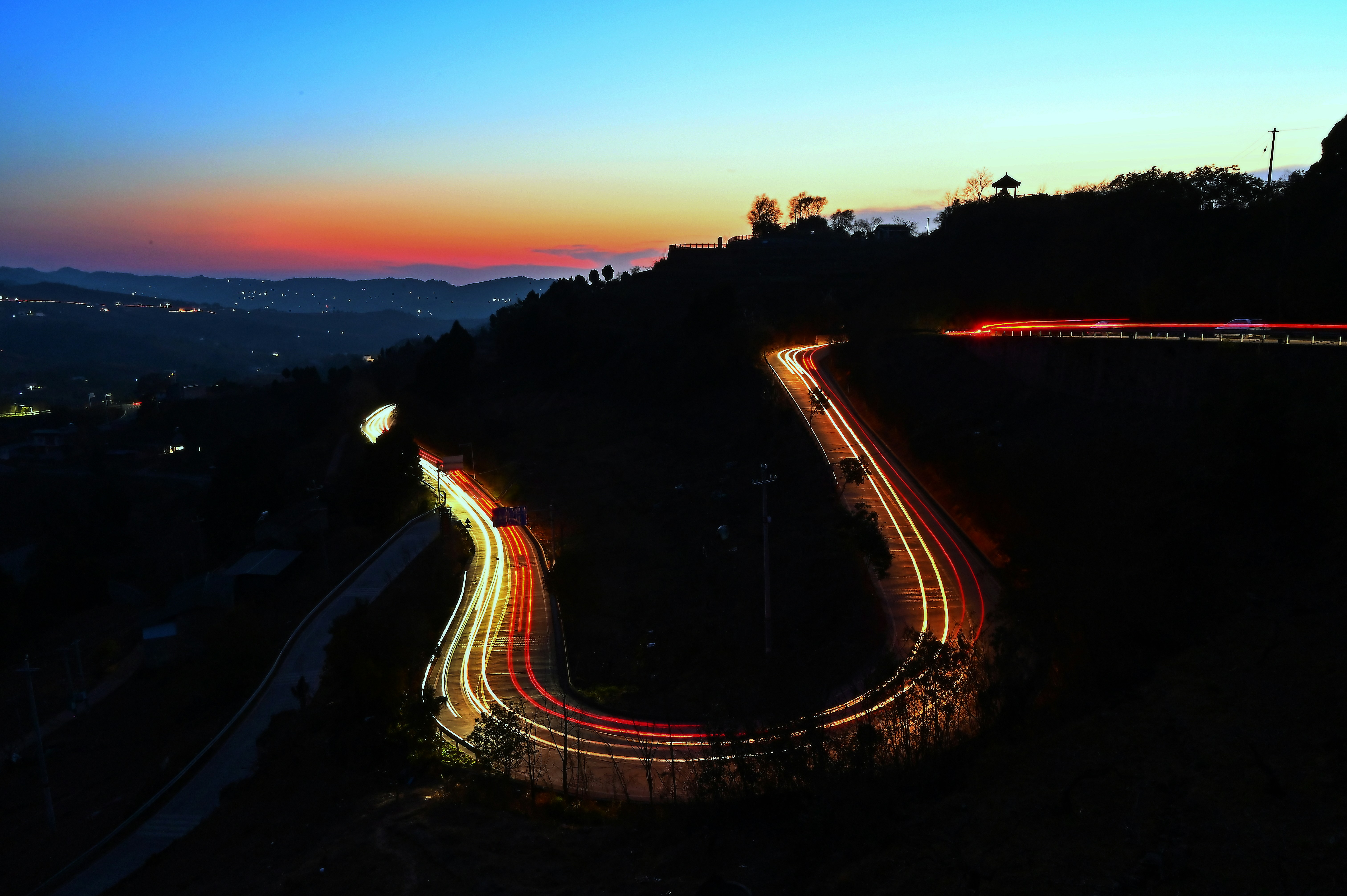 Twilight highway winds down a hillside, car trails creating curving ribbons of light against dark terrain; distant city lights glow on the horizon.