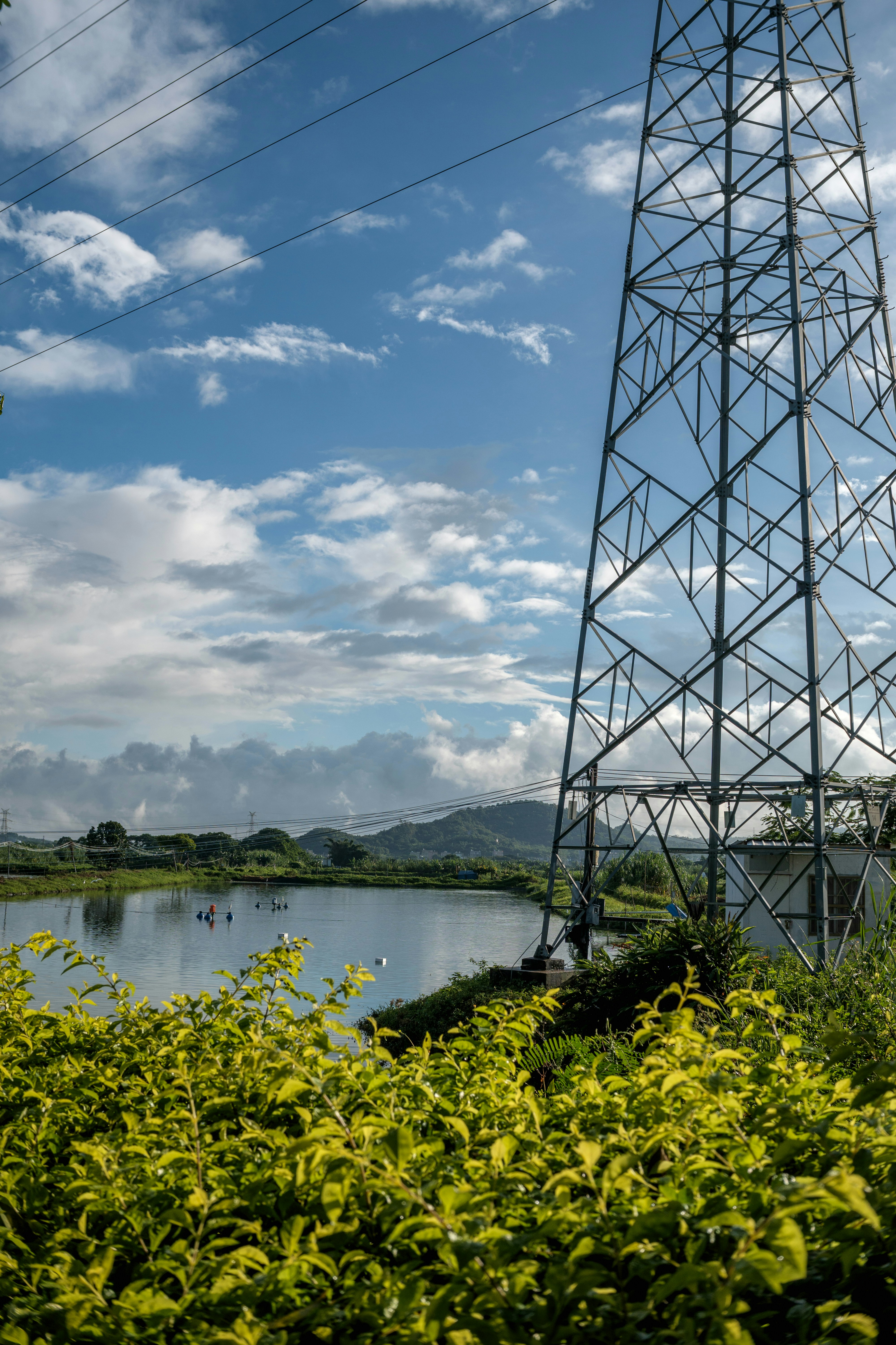 A metal lattice transmission tower rises on the right beside a calm lake with a green foreground and distant hills under a bright blue sky.
