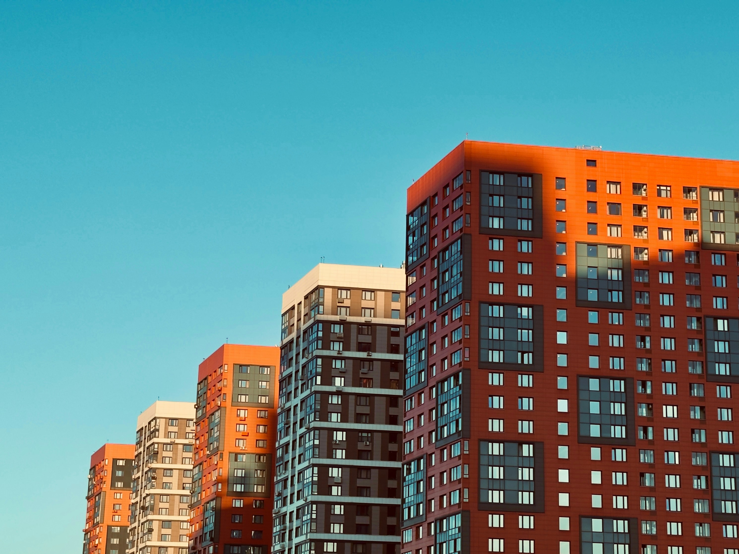 City skyscrapers with orange-brick facades reach toward a vivid blue sky, clean lines and stacked windows defining the modern urban panorama.