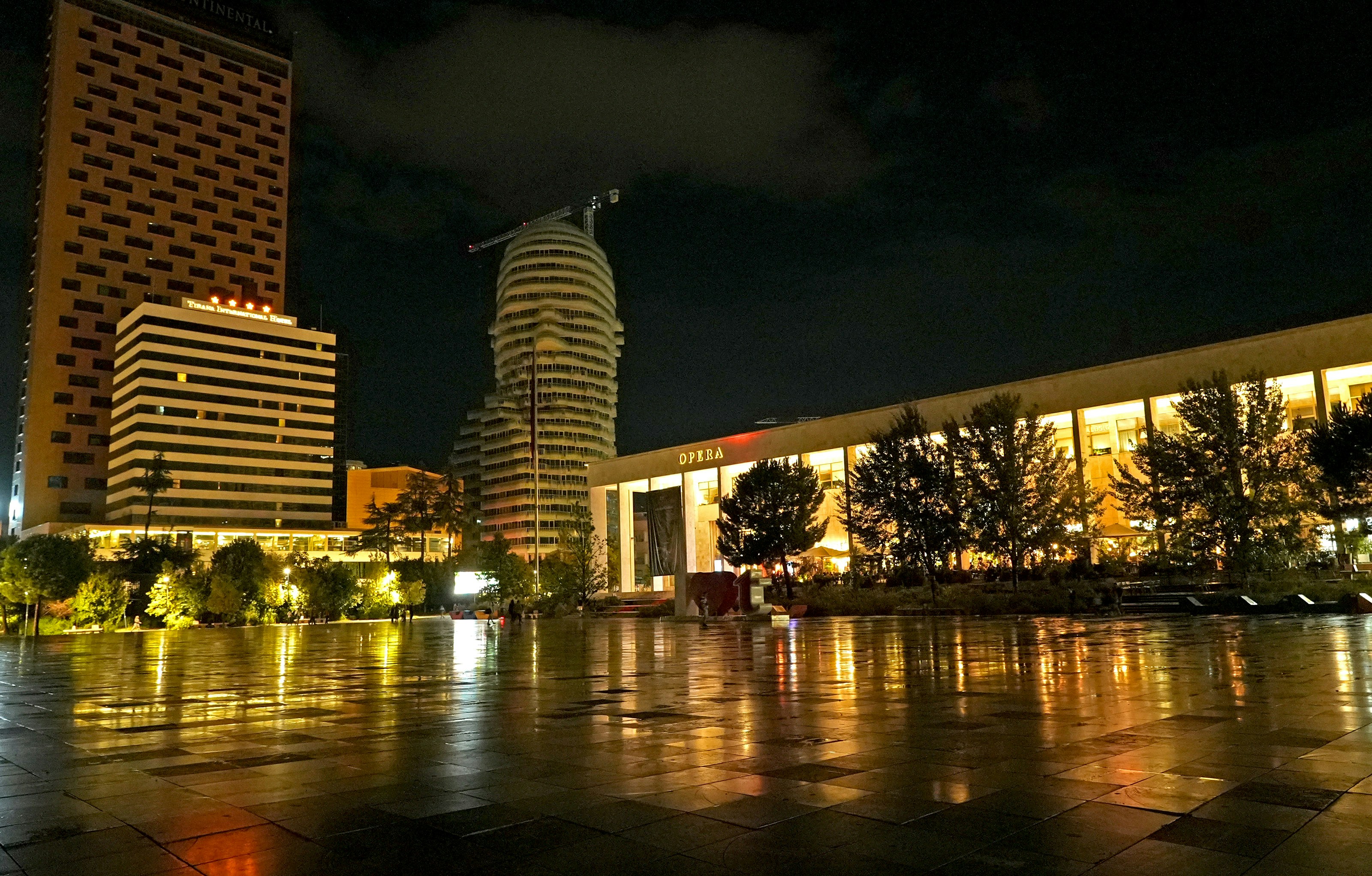 Night scene of a modern city plaza with illuminated buildings and reflecting wet pavement, foregrounds a broad, reflective surface and a cluster of trees along a lit facade. A logo-like building front anchors the composition.