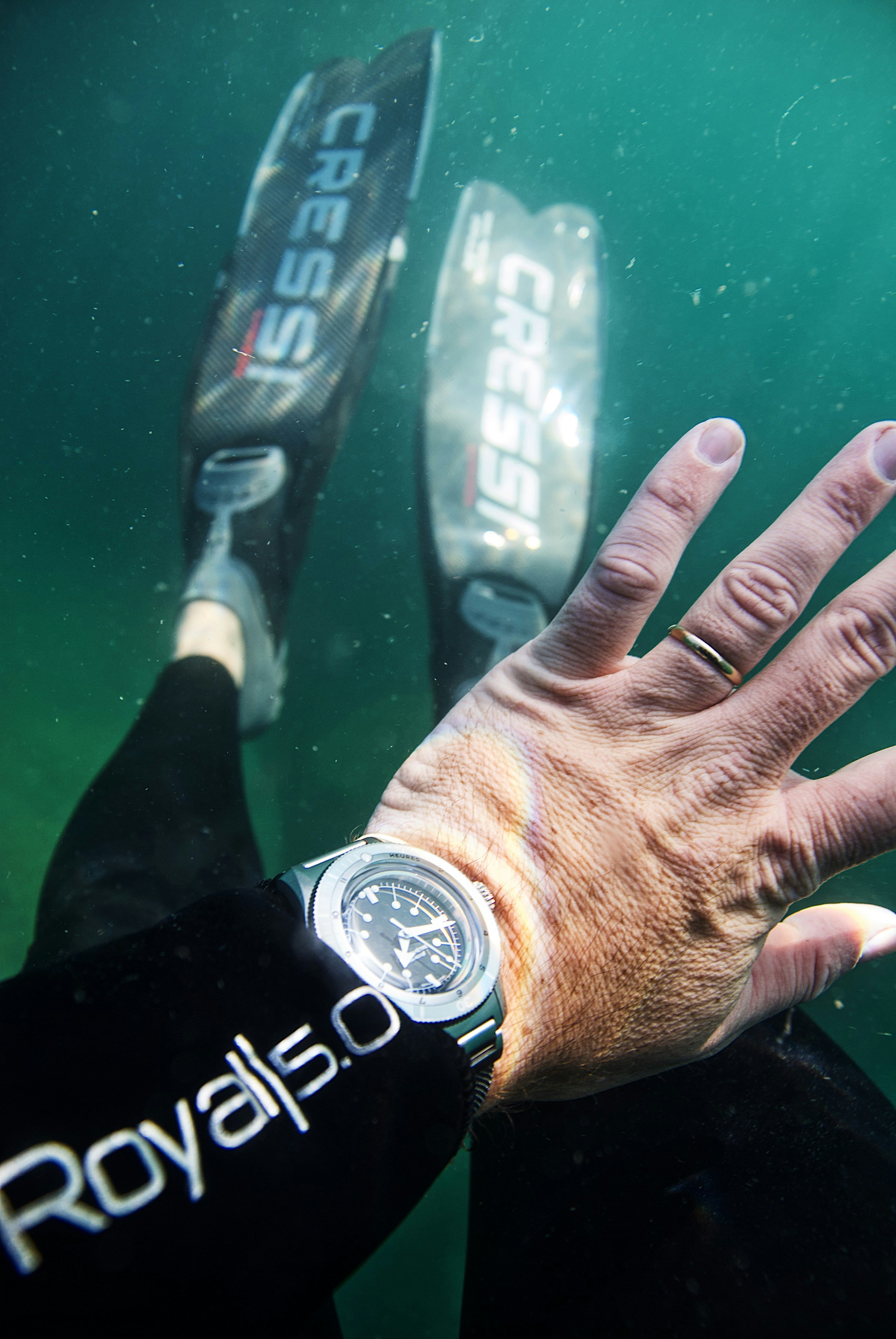 Diver's wristwatch and hand with Royal50 branding visible underwater, flippers and water droplets in view for context and scale.