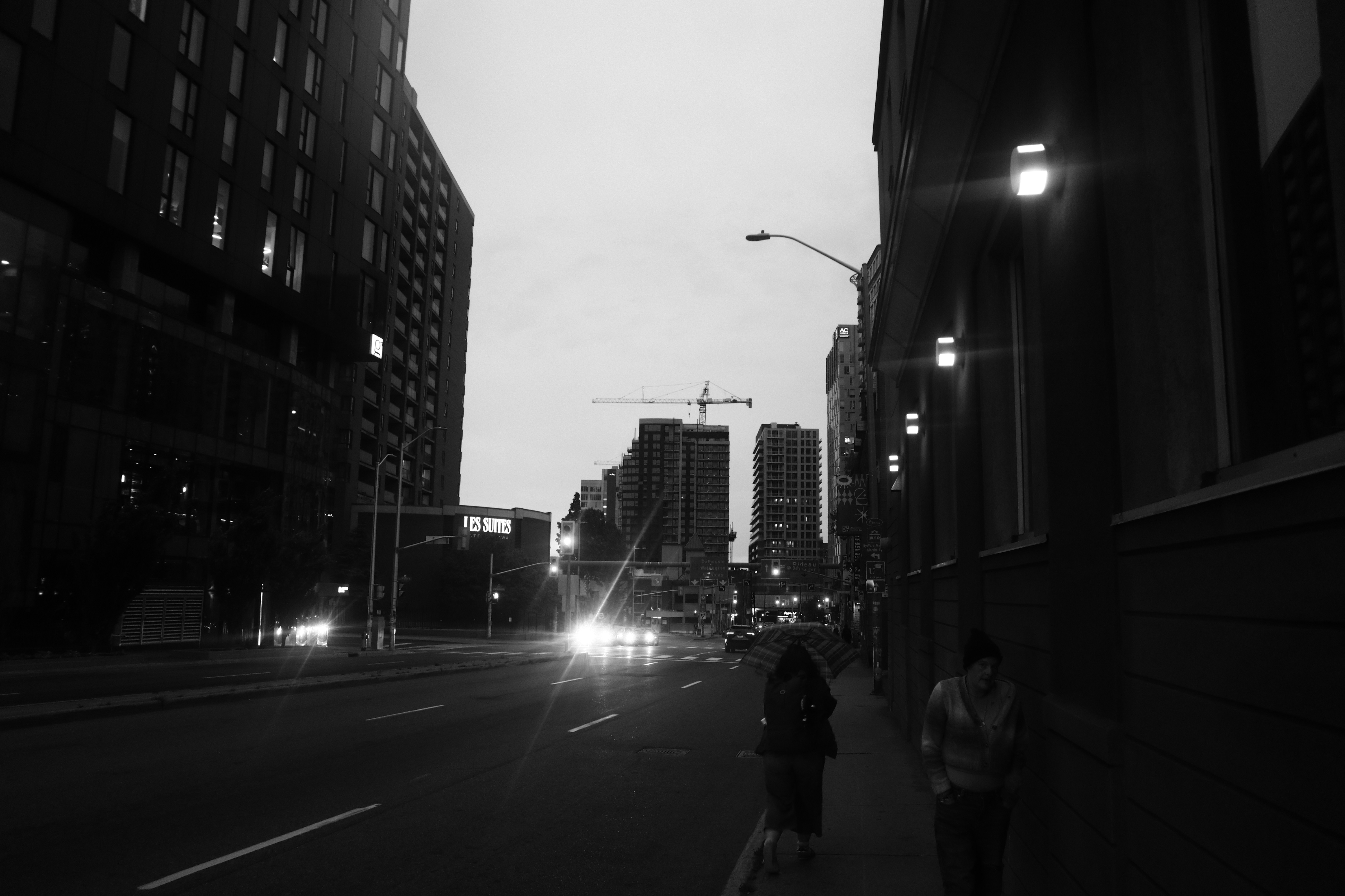High-contrast nighttime city street scene with two pedestrians along a building wall and traffic in the distance; monochrome emphasis on urban lighting and perspective.