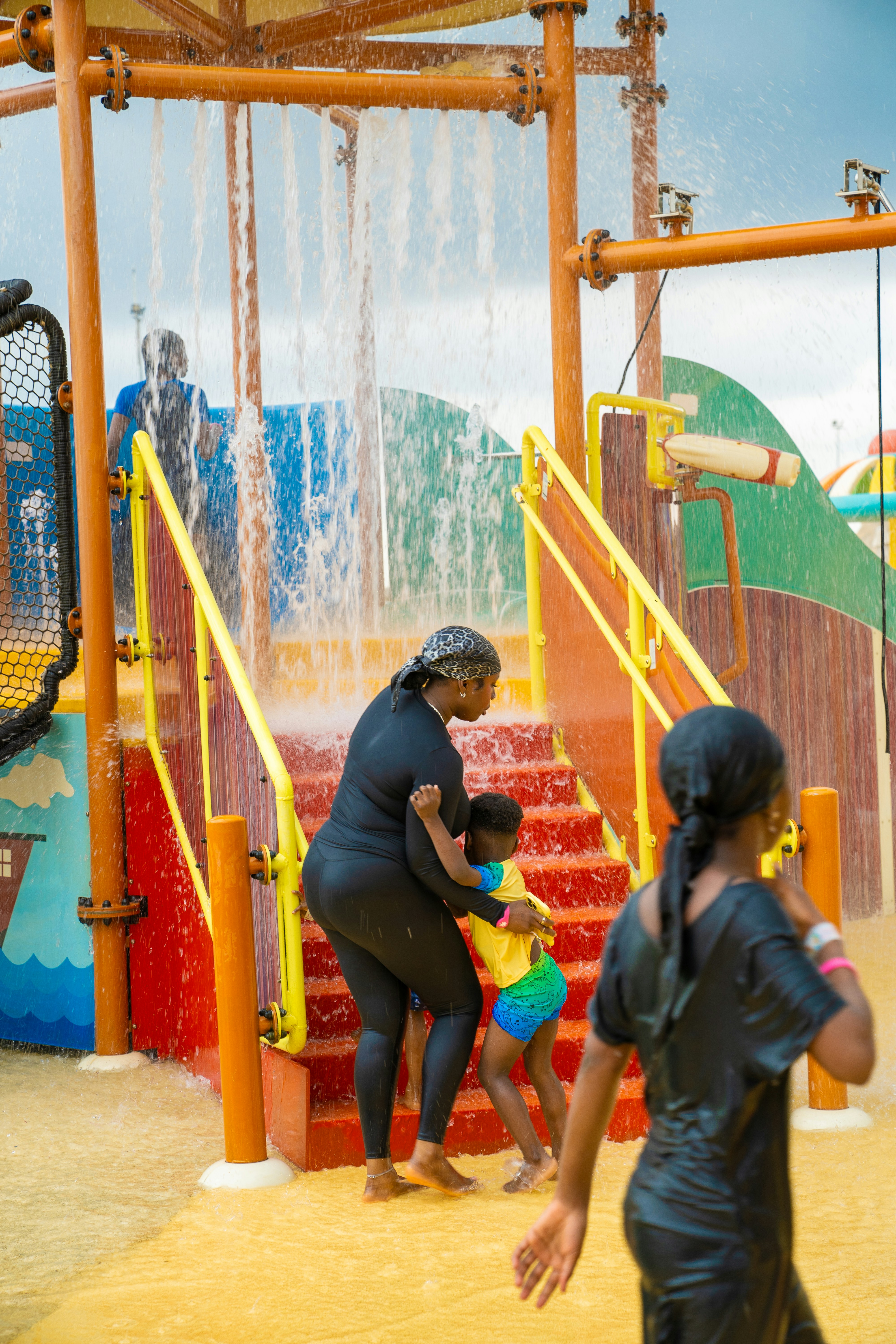 A mother in a black wetsuit steadies a young child at a colorful water playground under a cascade of jets, while another woman in the foreground watches. The scene captures playful activity and summer fun at a splash facility.