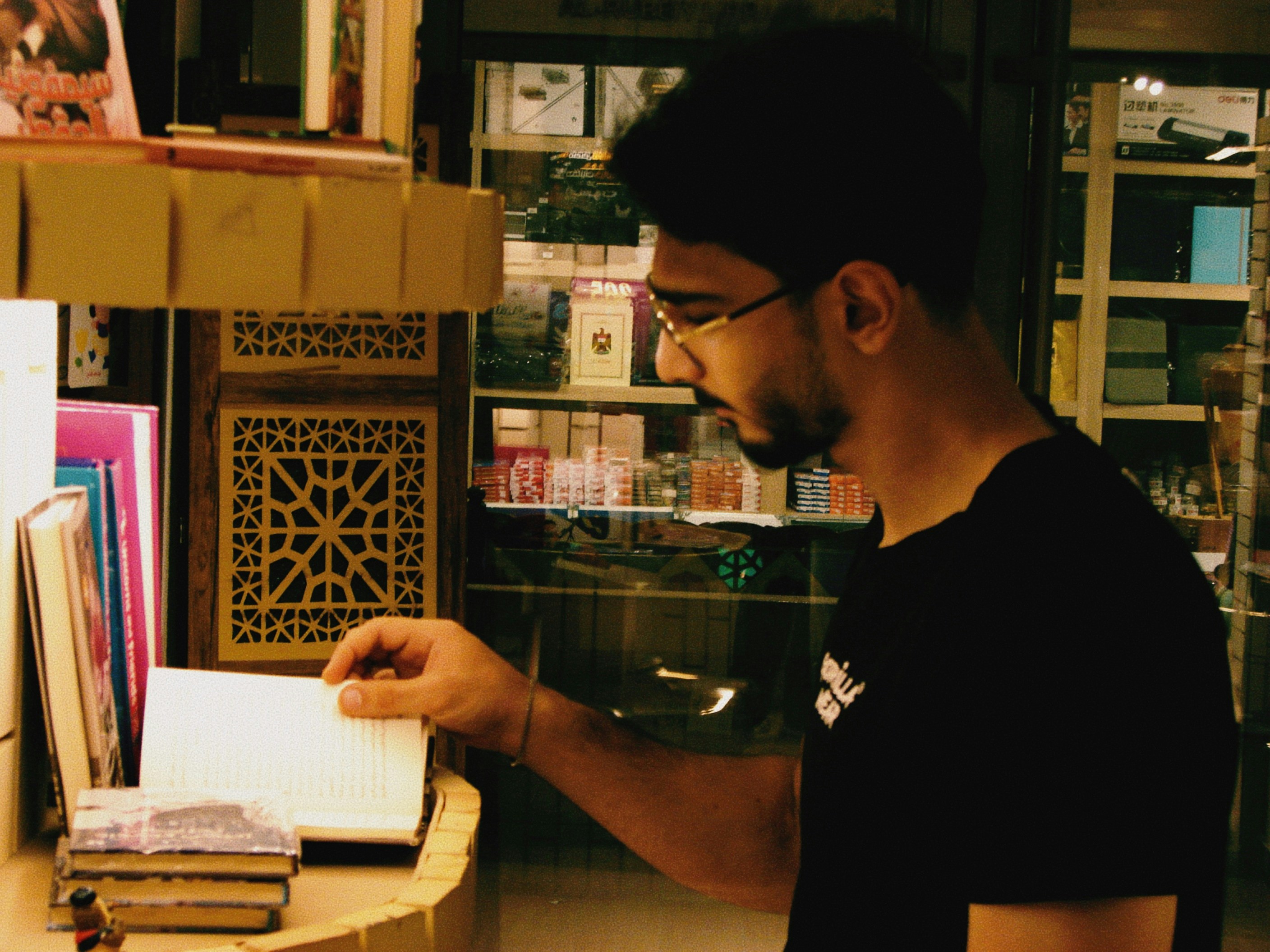 Man with glasses in a bookstore examines an open notebook among stacked shelves and books in warm light. This is a candid shot capturing a quiet reading moment in a shop.