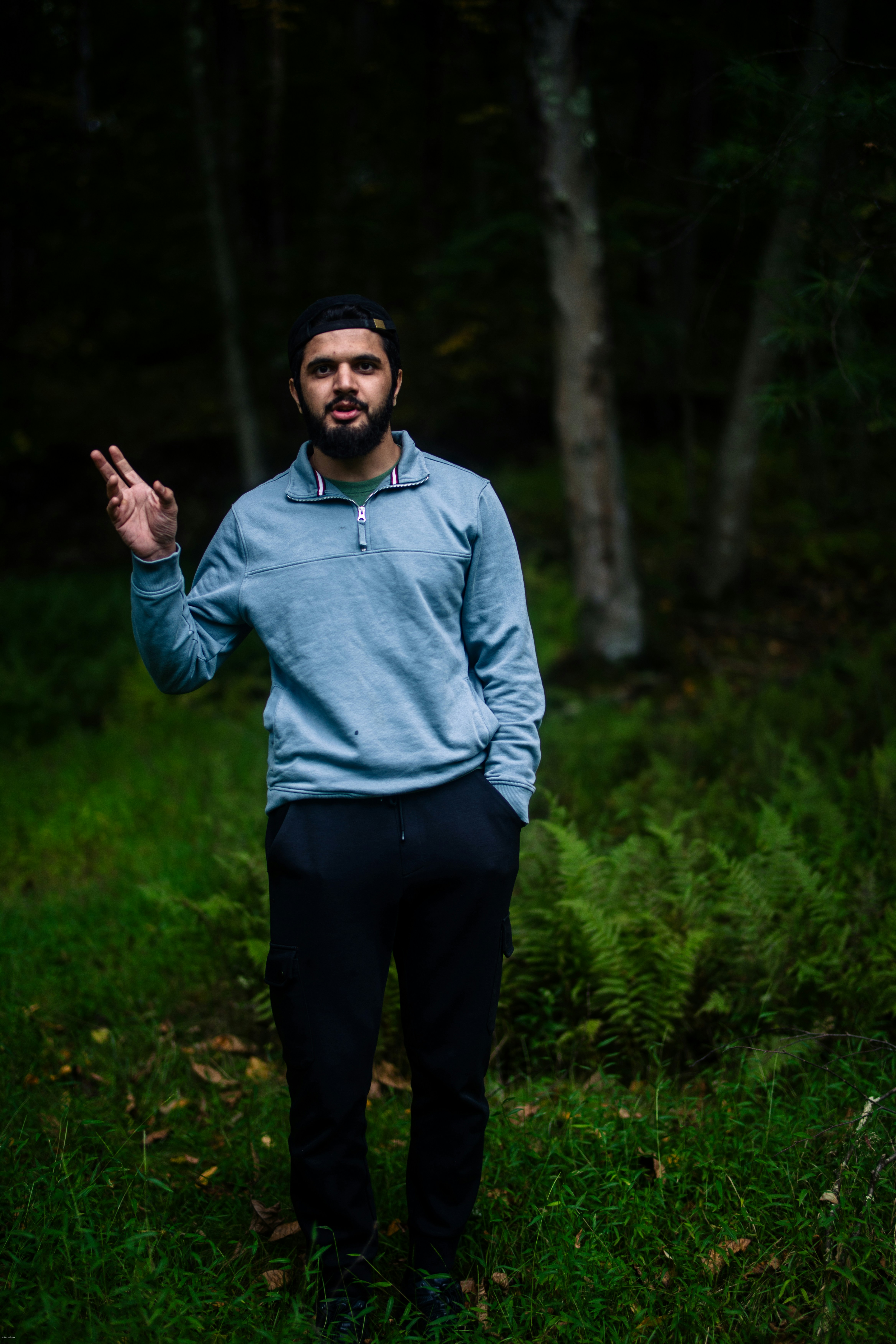 A man stands in a forest clearing, one hand raised in a casual wave while wearing a light-blue pullover and dark joggers, with dense greenery and trees in the background. Photograph capturing a relaxed outdoor moment.