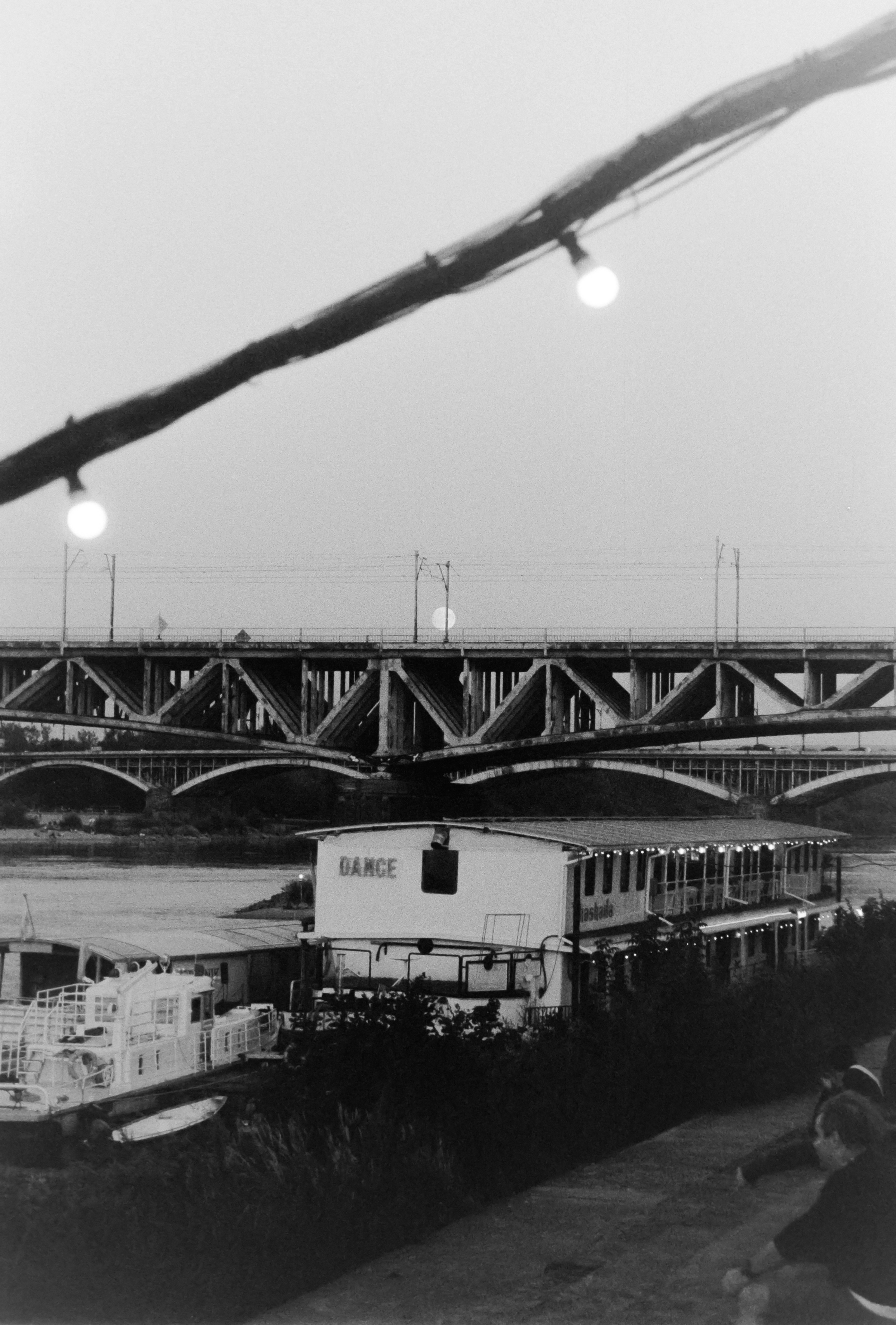 A black and white view of a river boat near a bridge with string lights