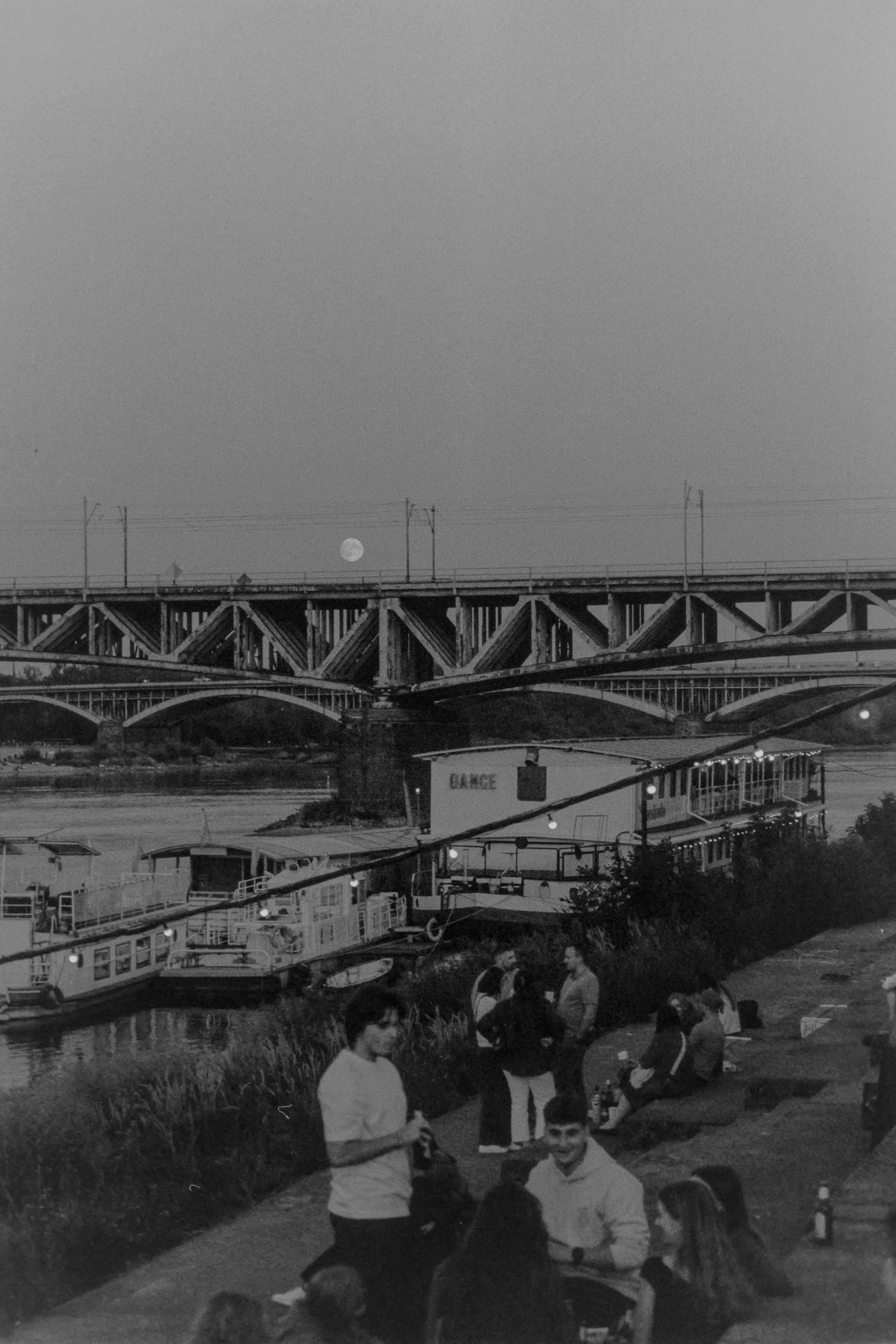 People gathered on a riverbank near moored boats beneath a large bridge at night