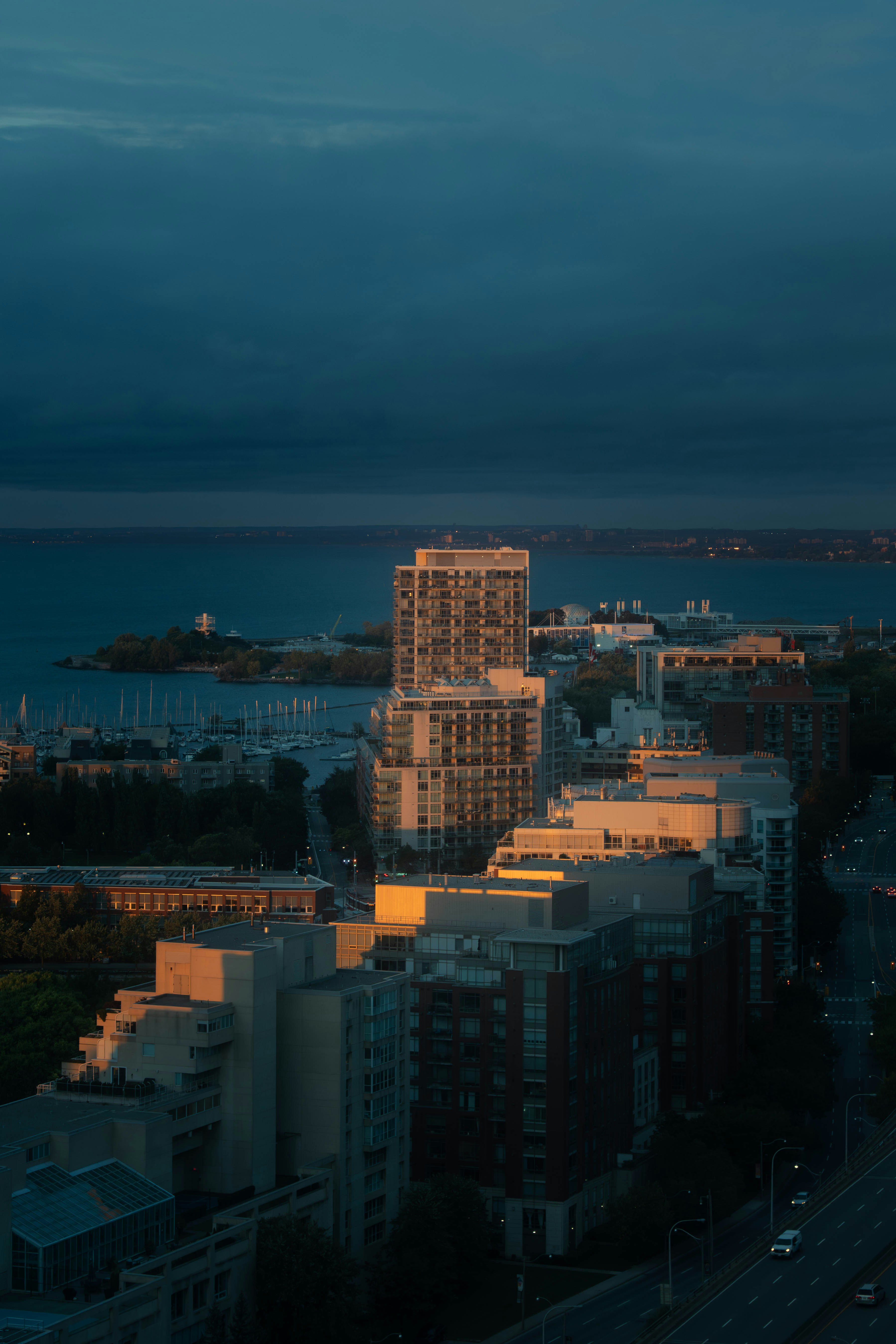 Urban harbor skyline with a cluster of modern buildings catching warm sunset light; foreground shows streets and residential blocks, while the sea and distant horizon sit under a moody sky.