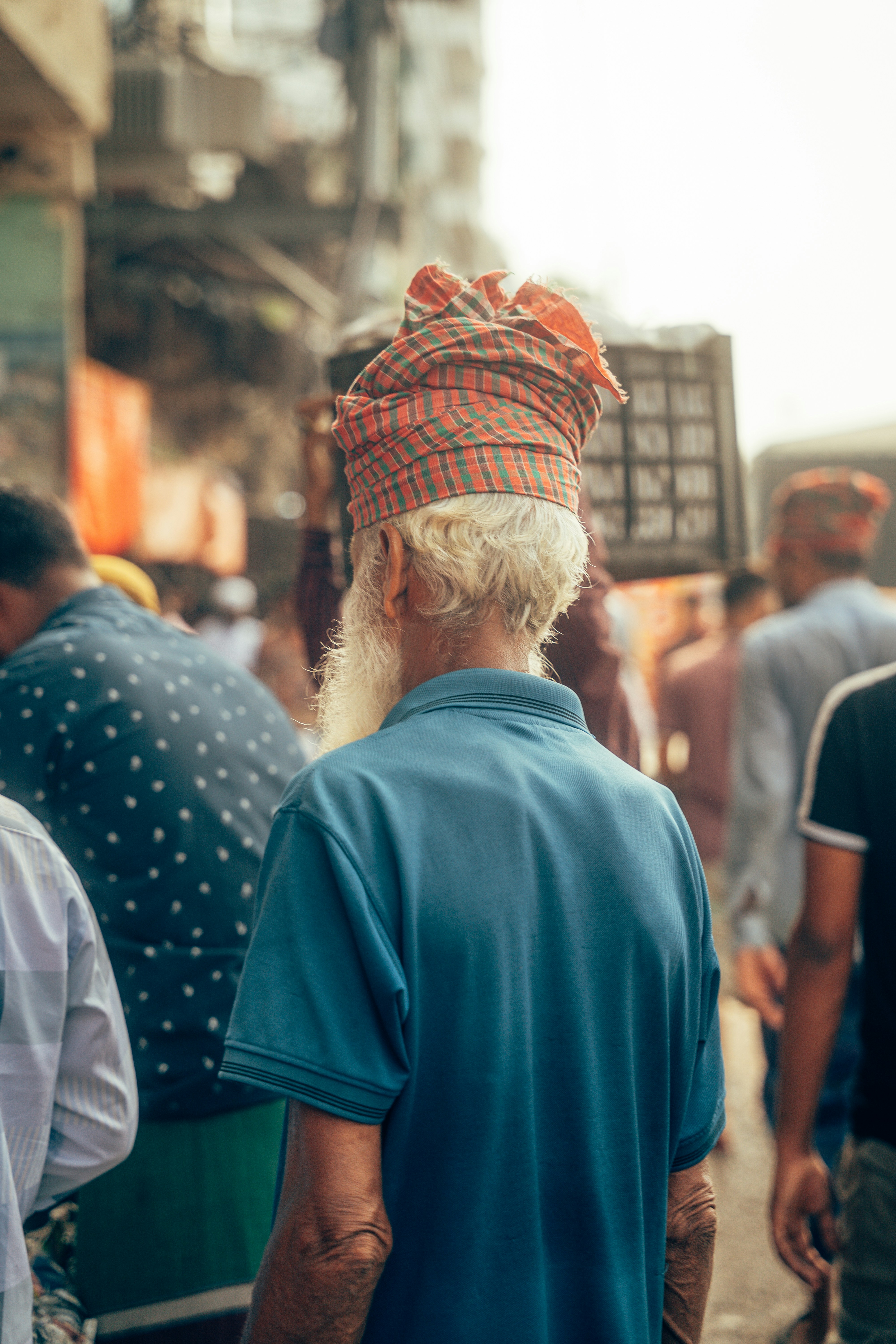A man with a red turban and blue polo stands among a crowded market scene, captured from behind to emphasize his distinctive headwear and the lively street around him.