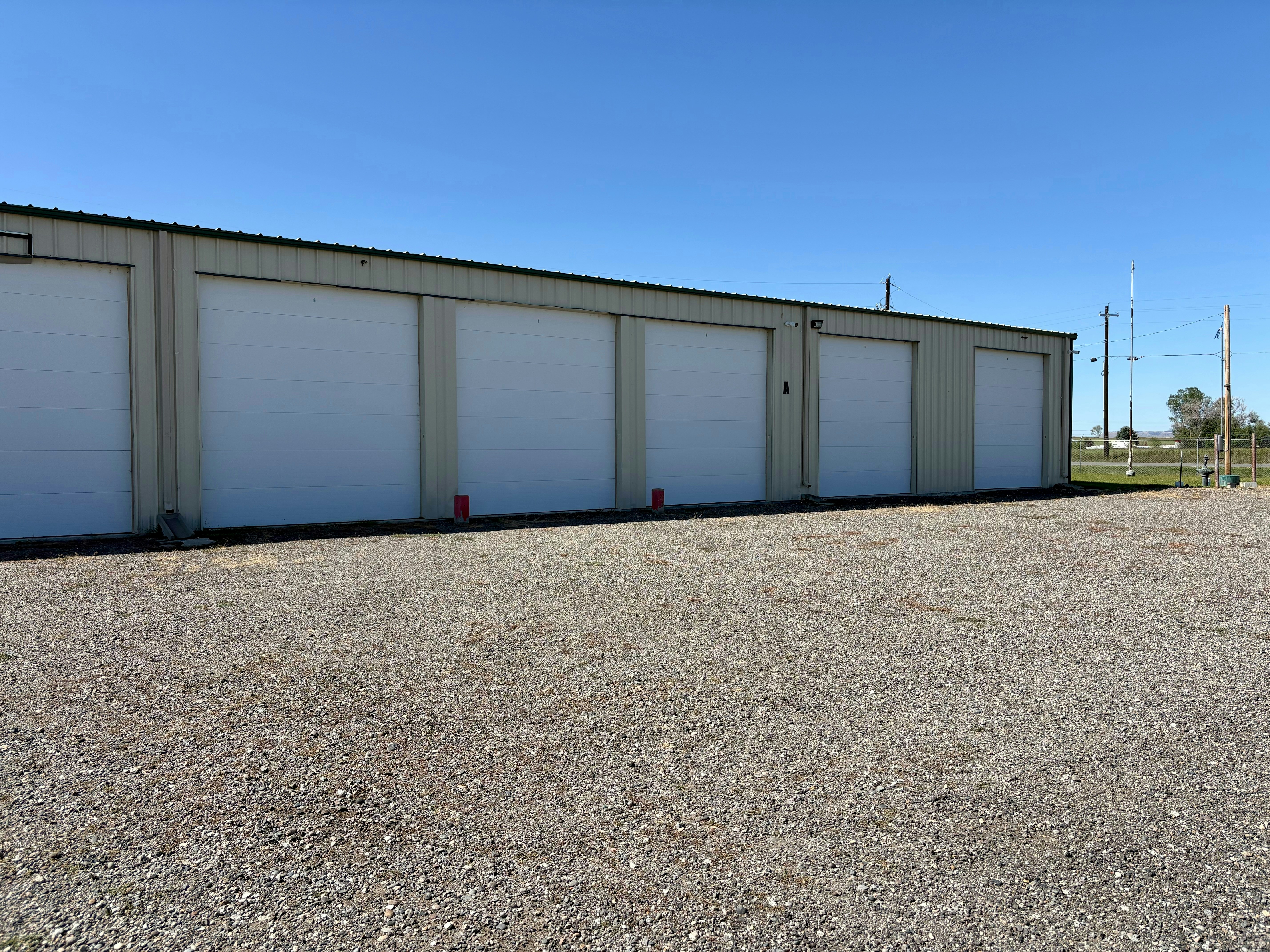 A row of white storage-unit doors sits along a gravel lot with a bright blue sky above; minimal foreground texture and a barren, utilitarian setting. Photograph shows a simple industrial exterior scene.