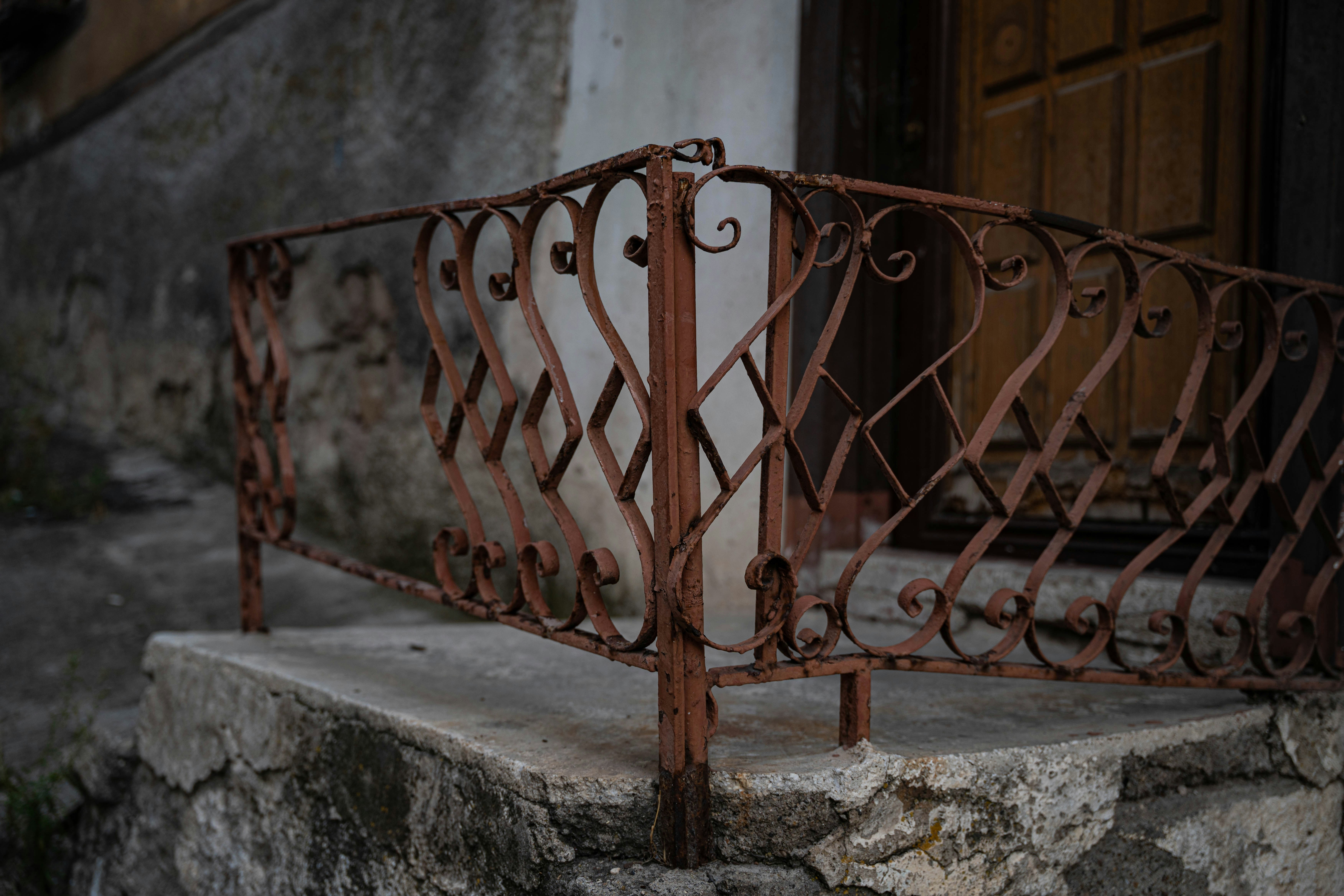 Rusty iron railing along a crumbling concrete step leads to a weathered doorway; muted tones and textured stone emphasize decay.