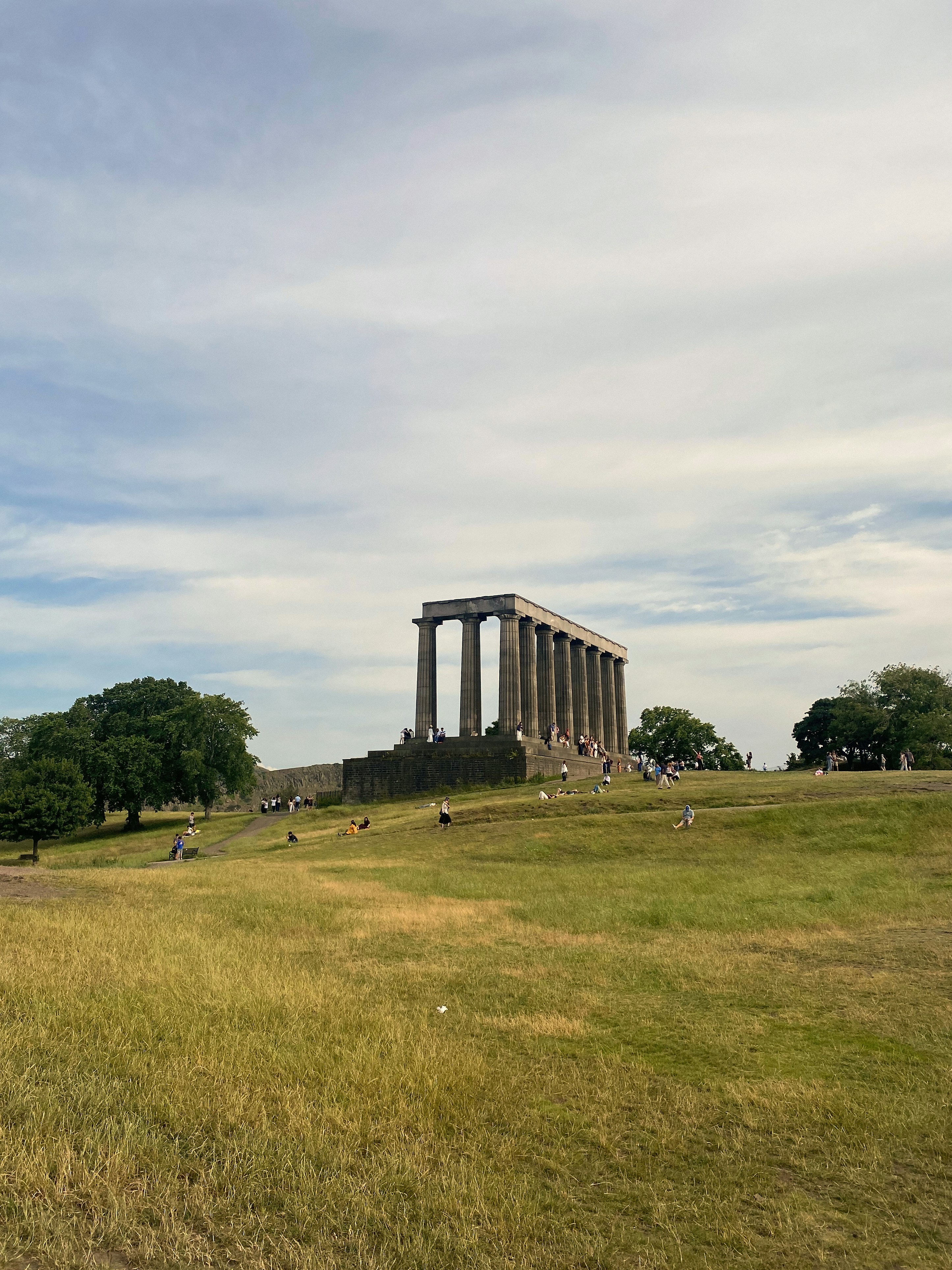 Stone ruins atop a grassy hill with visitors exploring and a wide, expansive sky in the background. A landscape-focused scene emphasizing the temple ruins as the focal point.