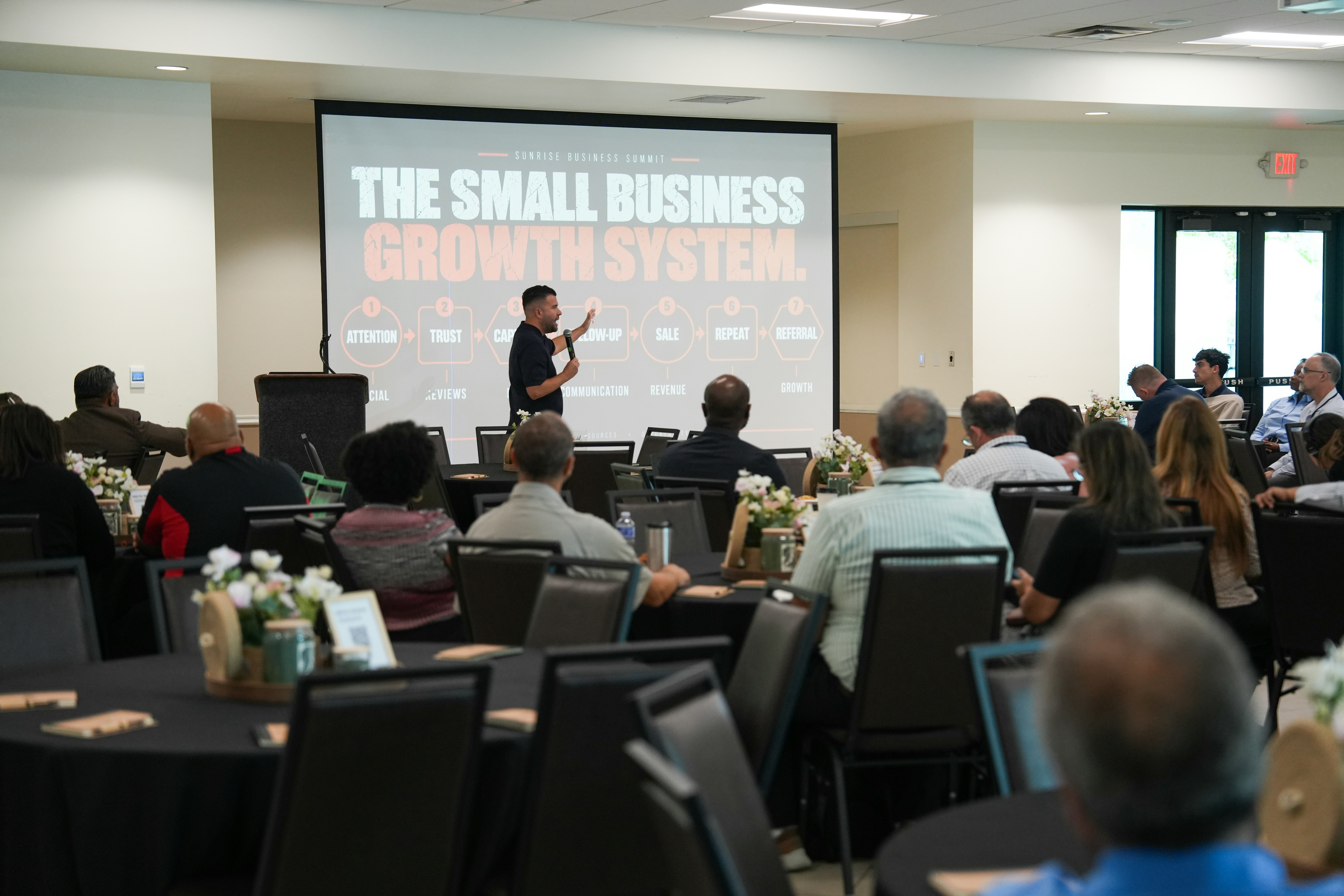 A speaker presents to a conference audience in a banquet-style room with a large screen displaying a business growth theme. The scene emphasizes a formal, educational event setup, including attendees and round tables.