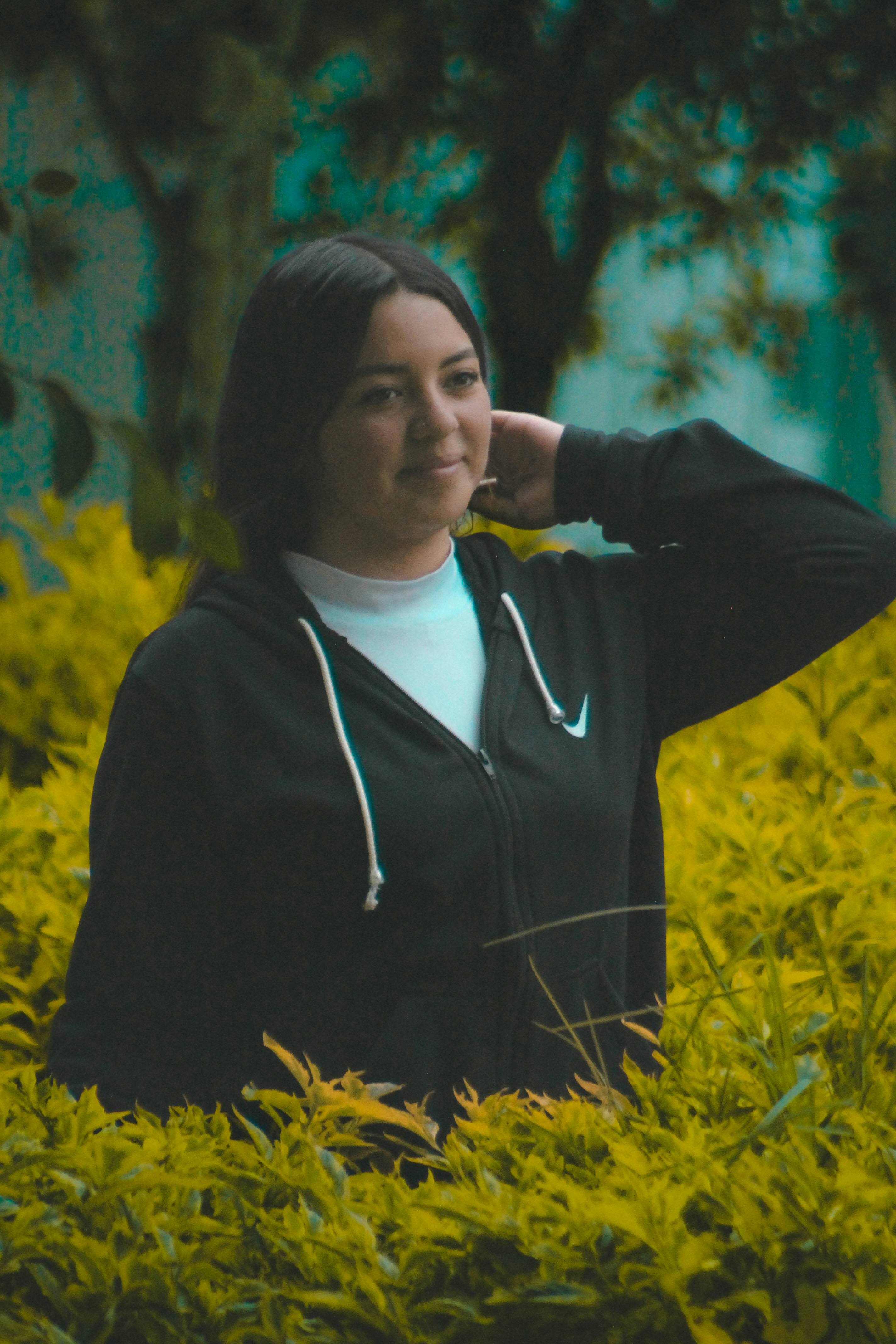 Portrait of a young woman in a black hoodie standing among yellow foliage, with a teal-toned background and a calm, introspective expression.