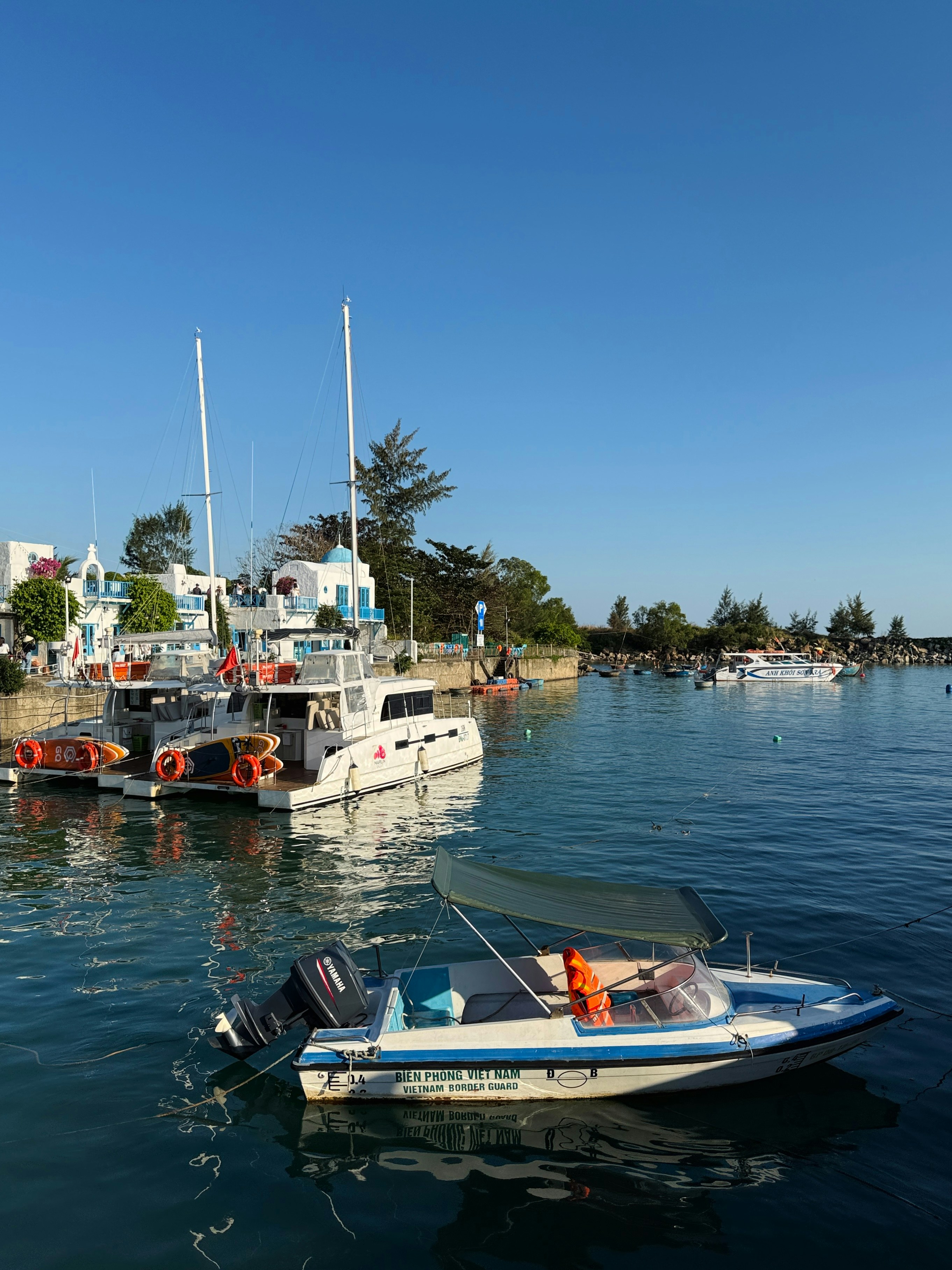Small motor boats rest on calm marina water with larger sailboats and a seaside village in the background.